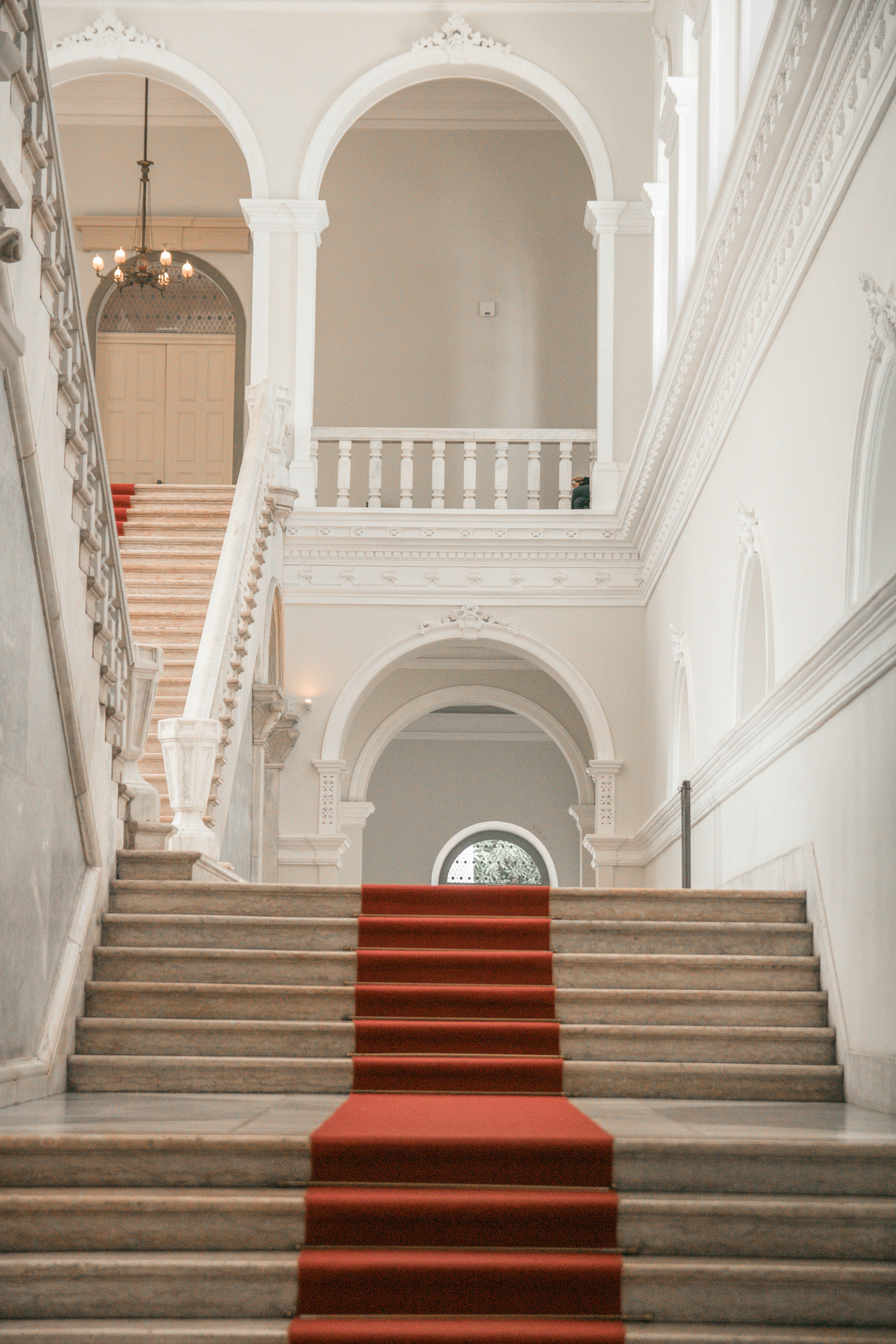 A grand staircase with a red carpet leads to ornate archways in a bright, white classical interior. A sense of symmetry and elevation dominates the composition, highlighting architectural detailing.