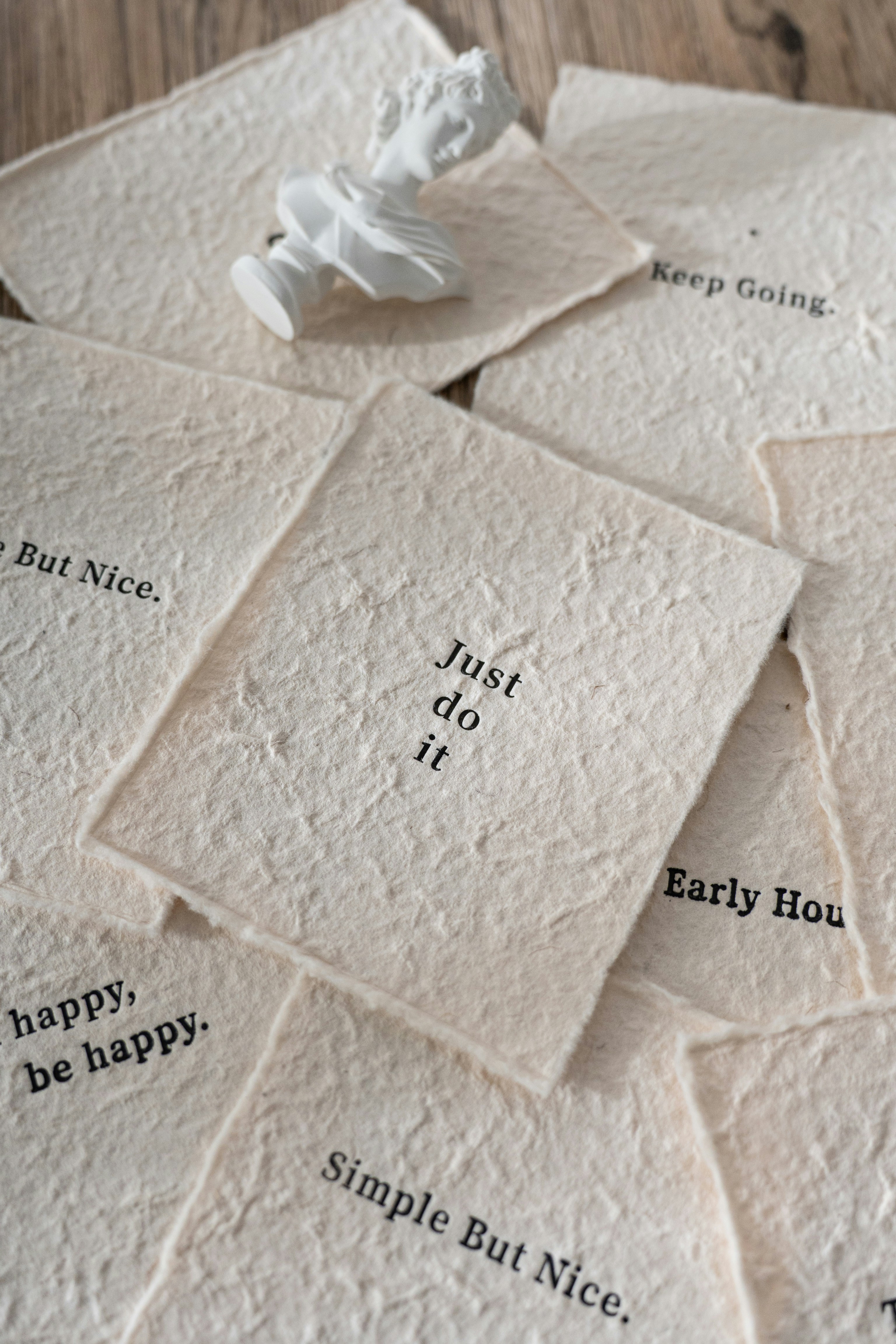Close-up of textured paper cards with motivational phrases arranged in a layered collage; the central card reads 'Just do it' in simple typography, conveying a minimal, tactile mood.