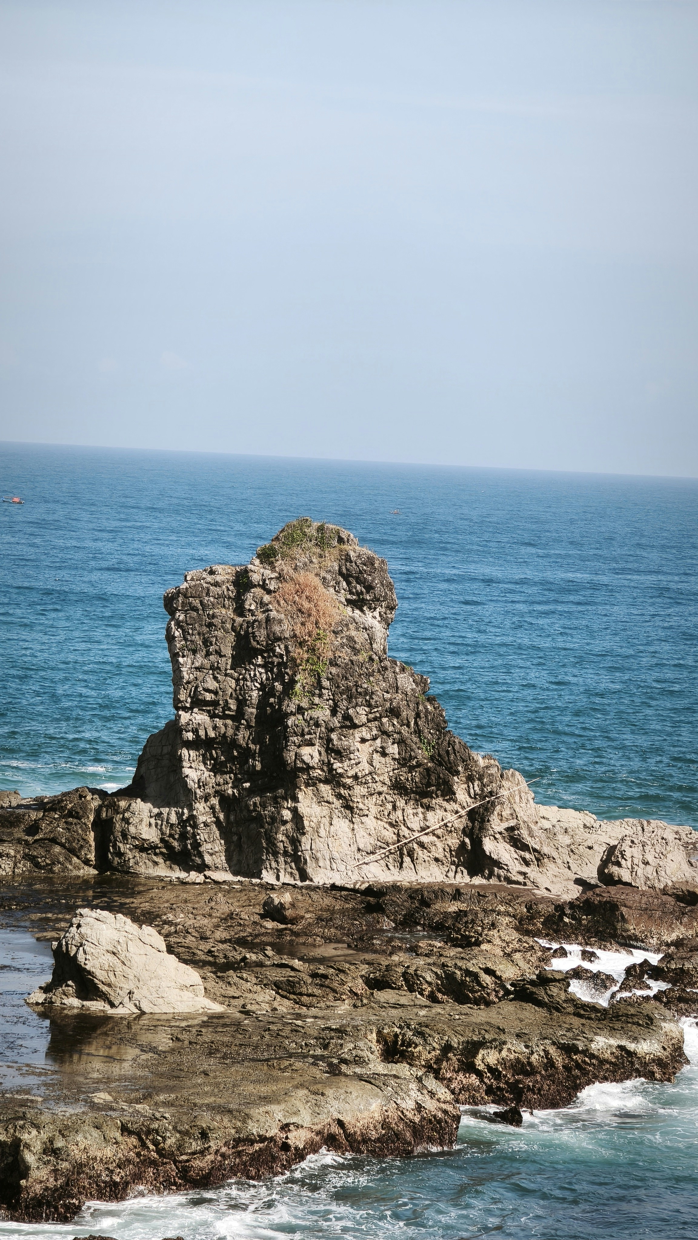 A lone rock formation rises from the rocky shore into calm blue sea, with distant horizon and clear sky above. The scene emphasizes coastal geology and sea spray textures.