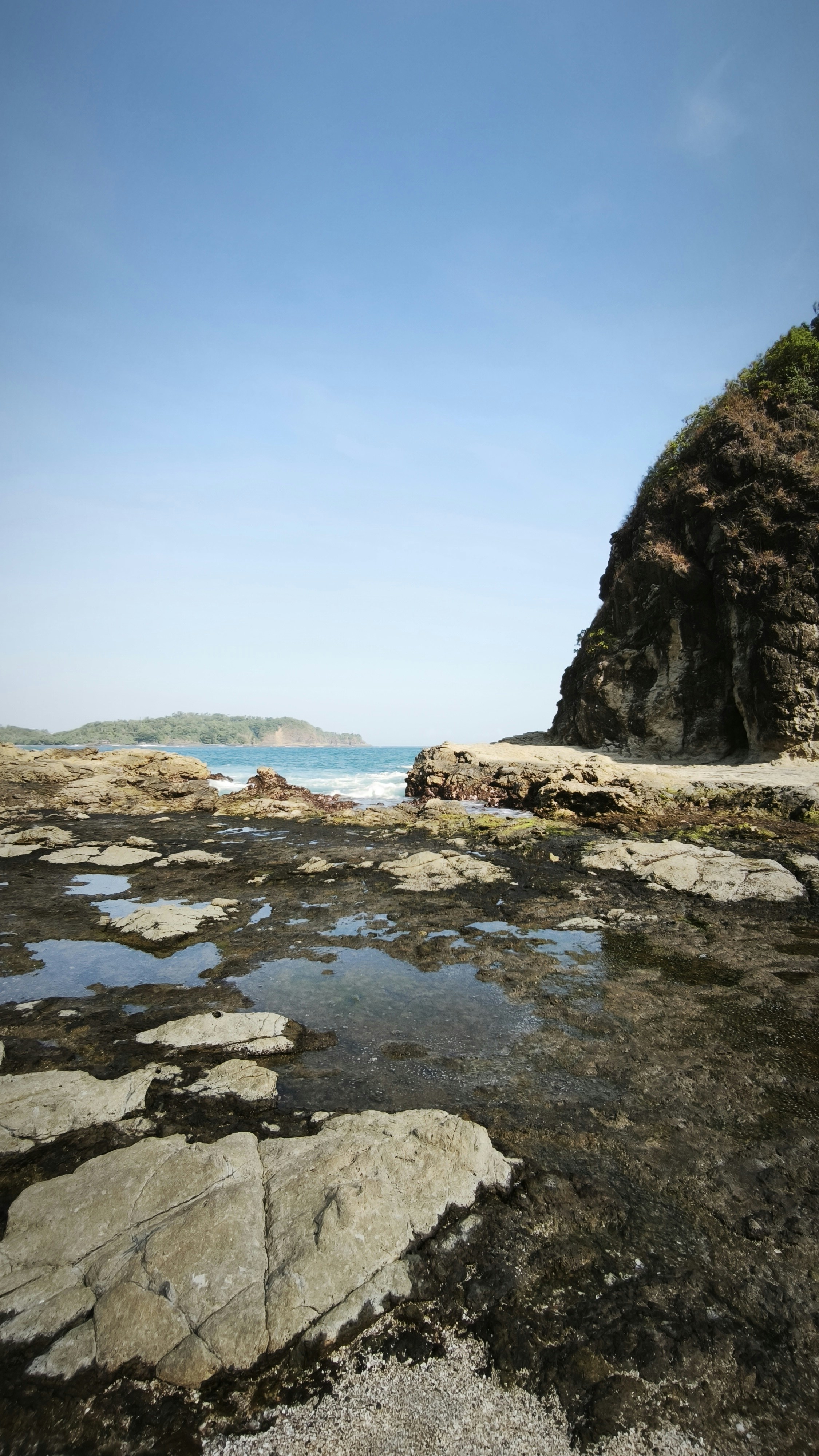 Rocky shoreline with tide pools stretching toward the blue sea and a towering cliff on the right, under a bright sky. A minimal landscape scene with natural textures and calm light.