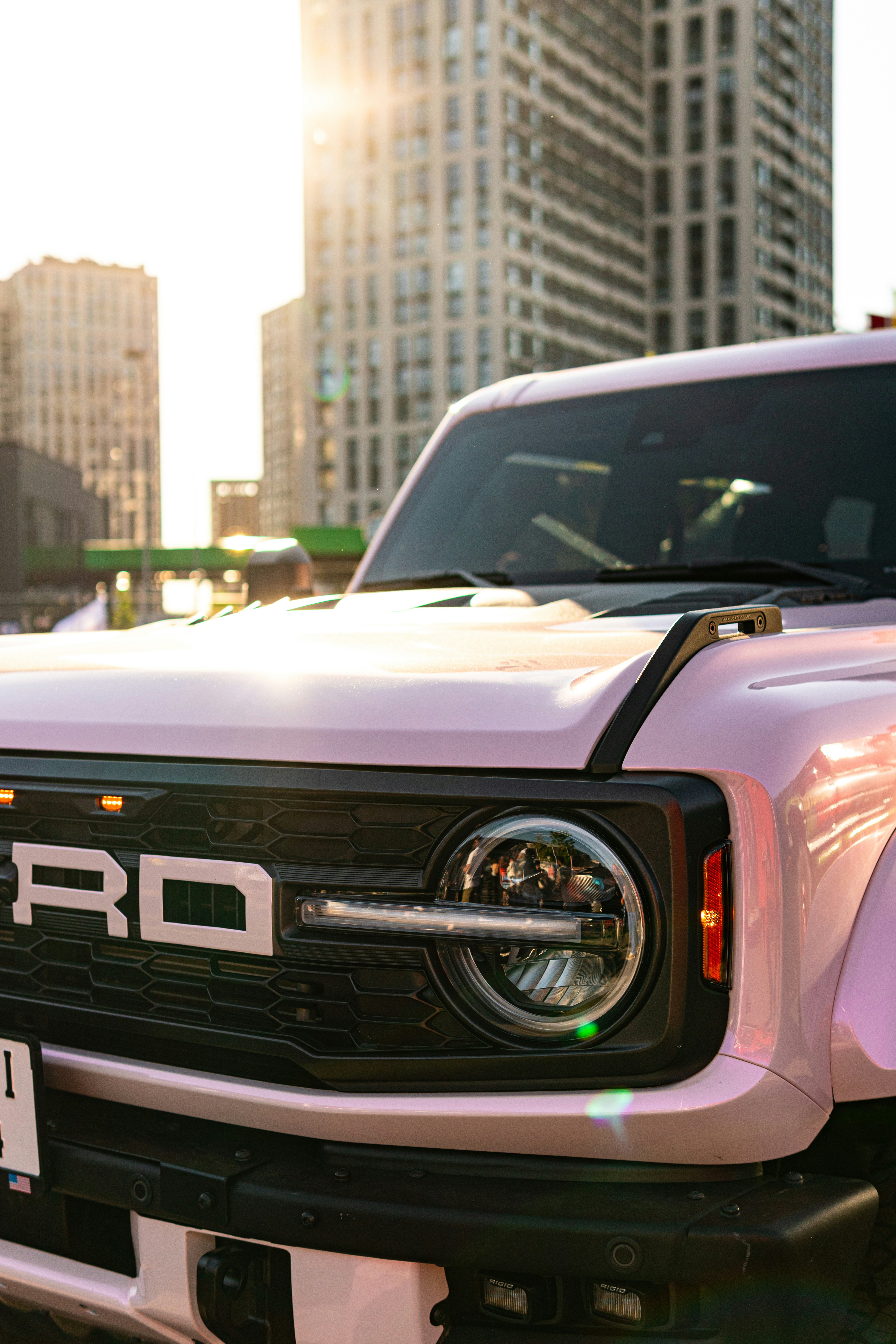 Close-up of the front end of a white Ford SUV with a round headlight and bold grille, set against tall city buildings and warm backlight.
