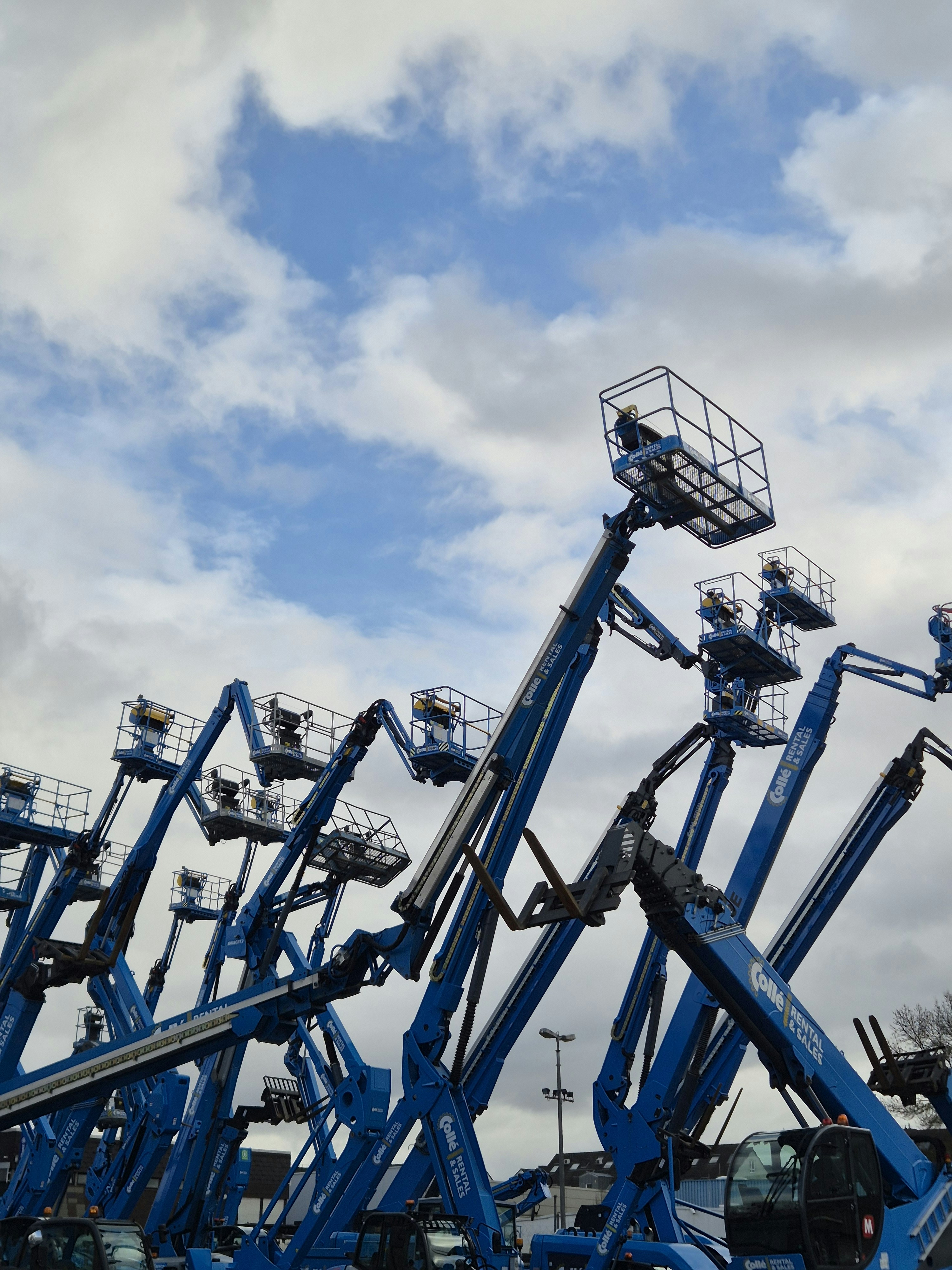 A dense field of blue industrial cherry-pickers stretches skyward under a cloudy day, their arms intertwining in a dramatic industrial landscape.