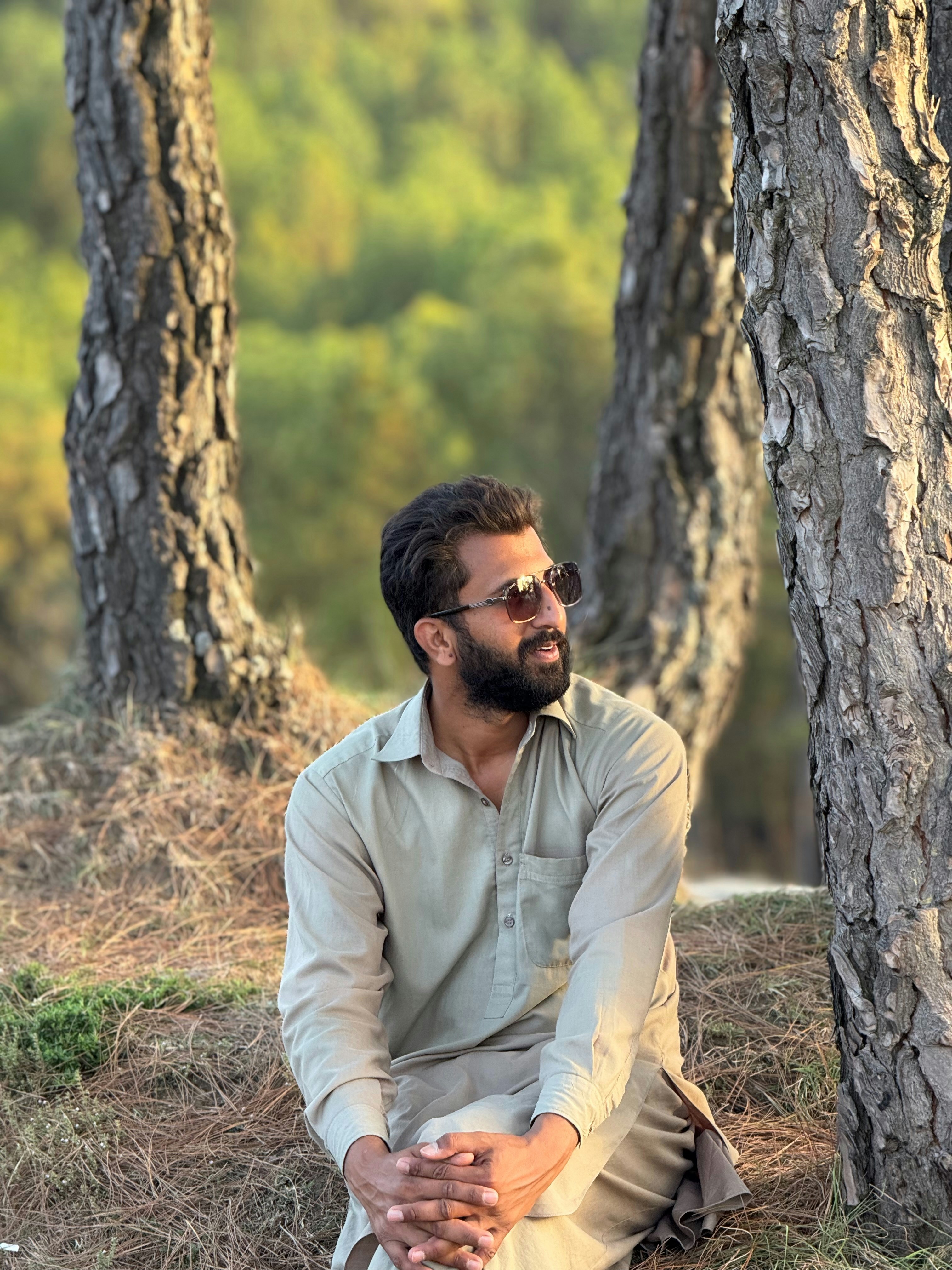 A bearded man in sunglasses sits among trees, looking right.