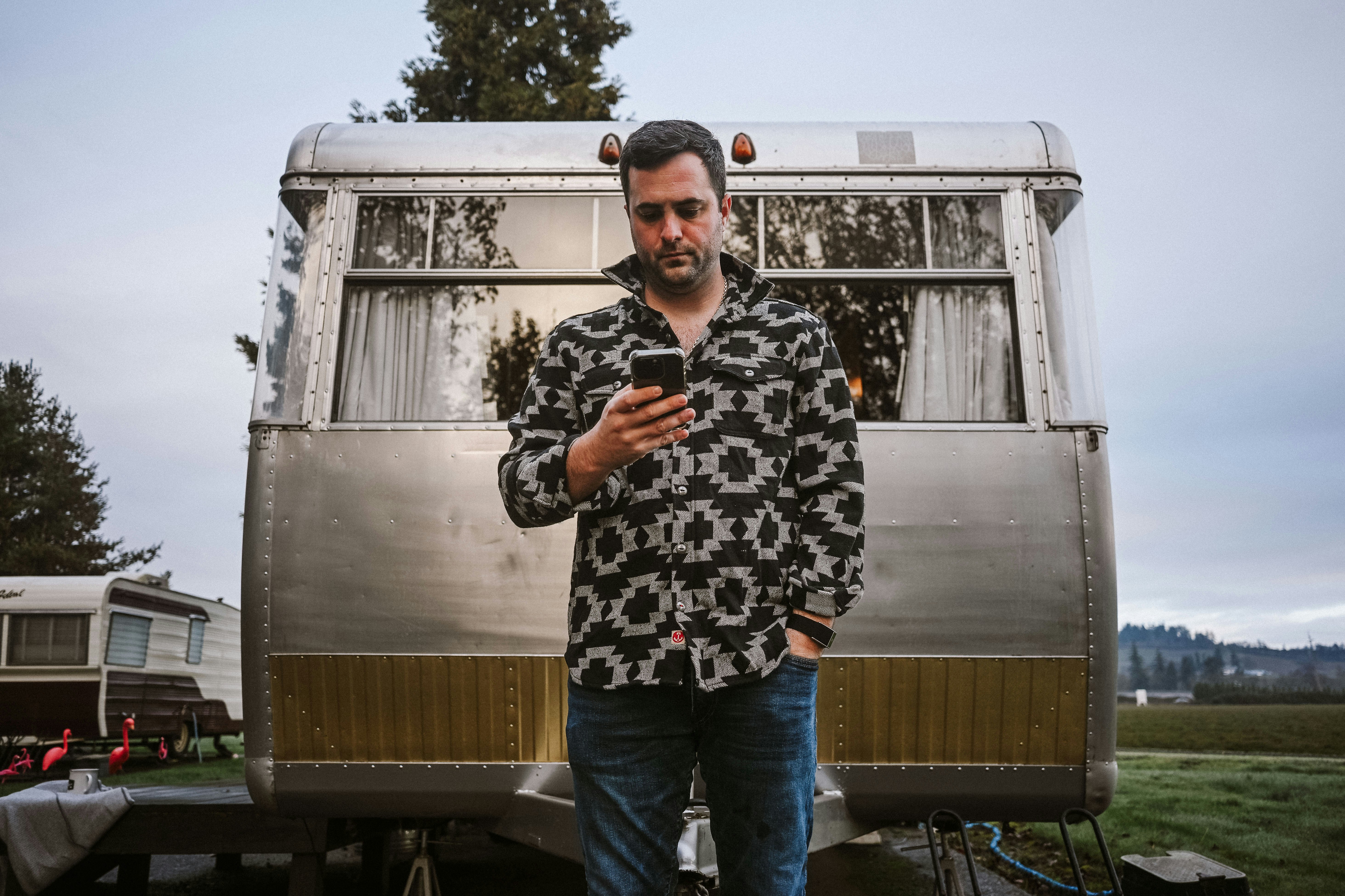 Man checks smartphone in front of a vintage travel trailer.