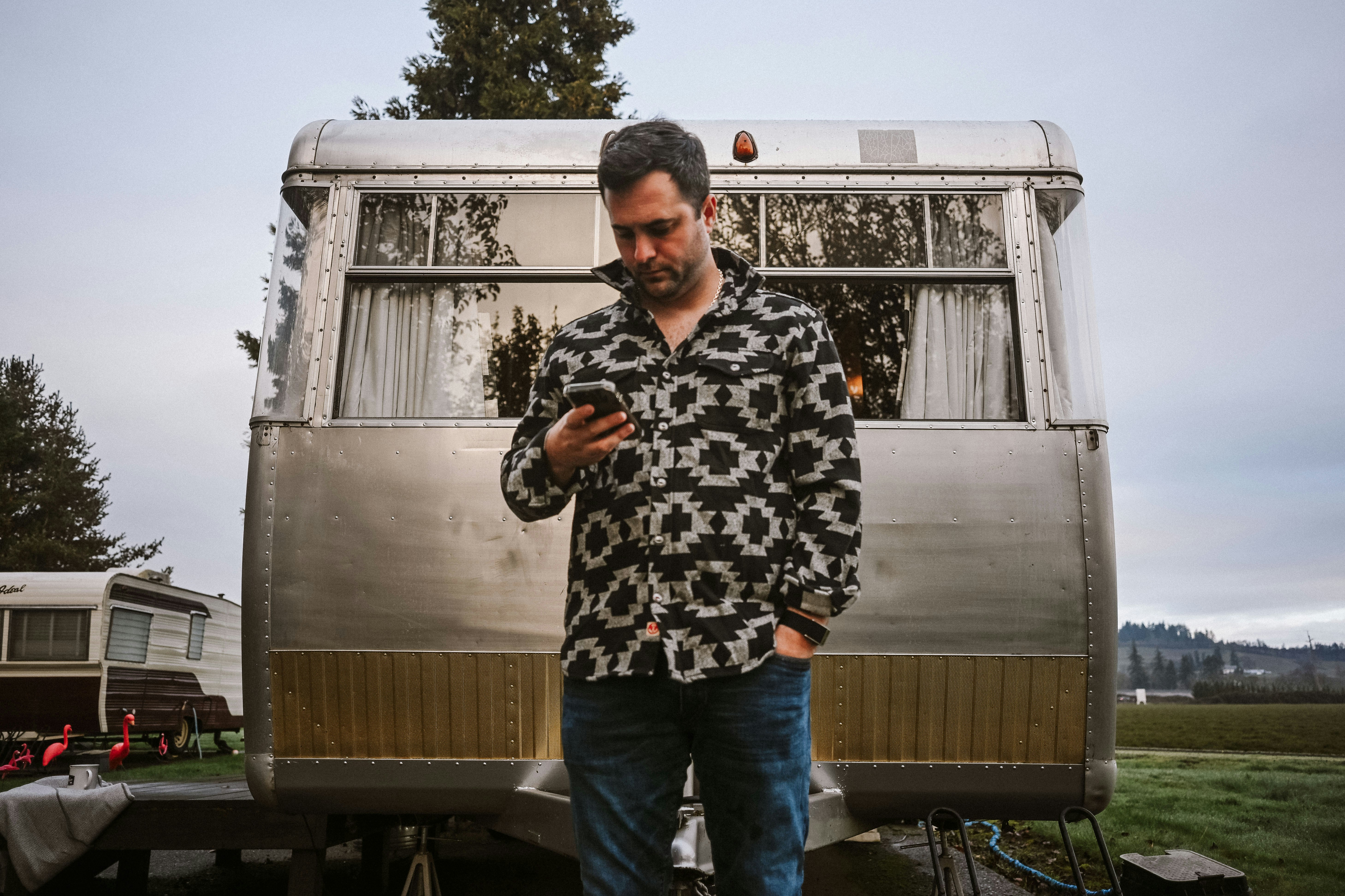 Man with patterned shirt absorbed in his phone in front of a silver trailer