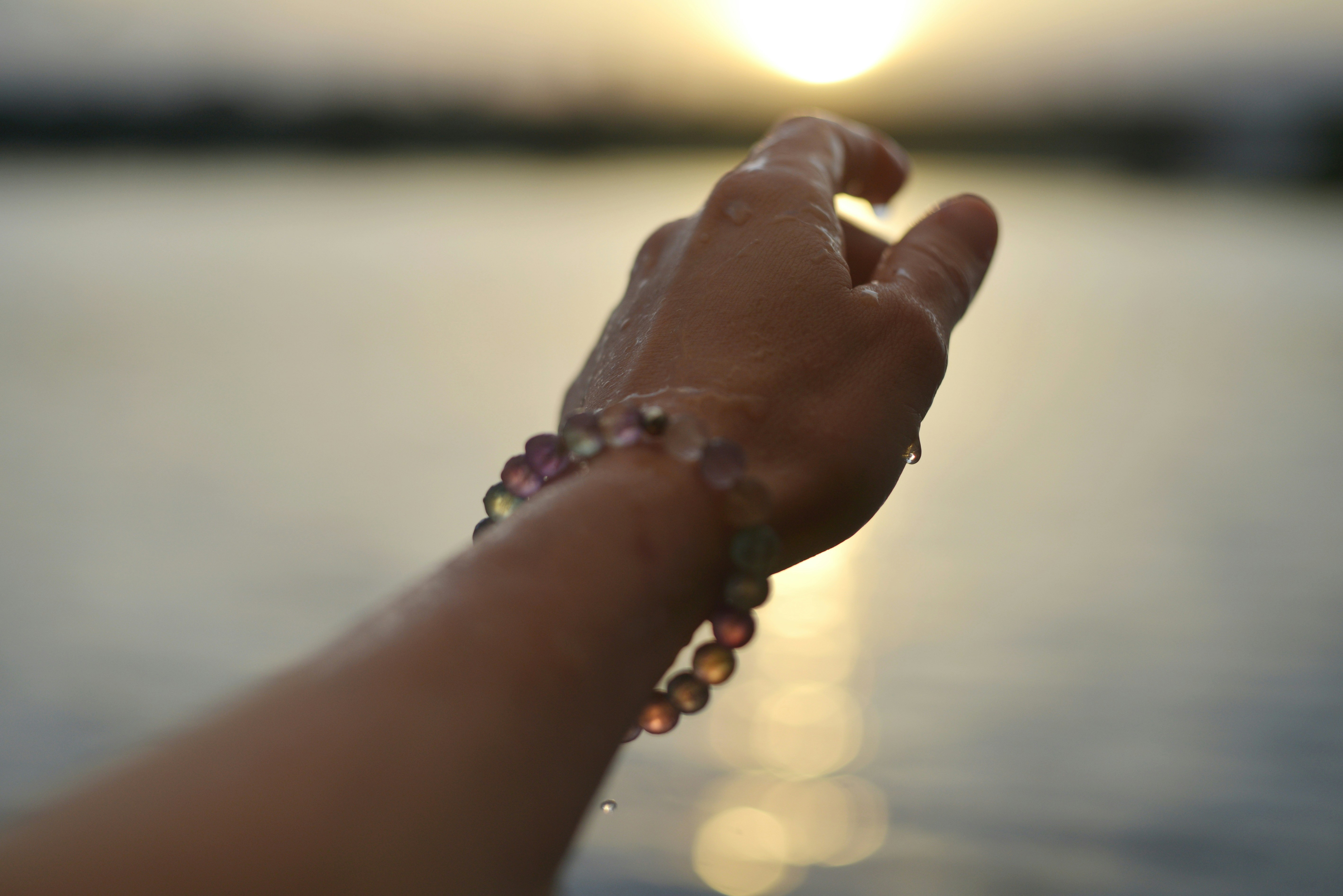 Beaded bracelet on an outstretched forearm reaches toward a sunlit water scene at dusk.