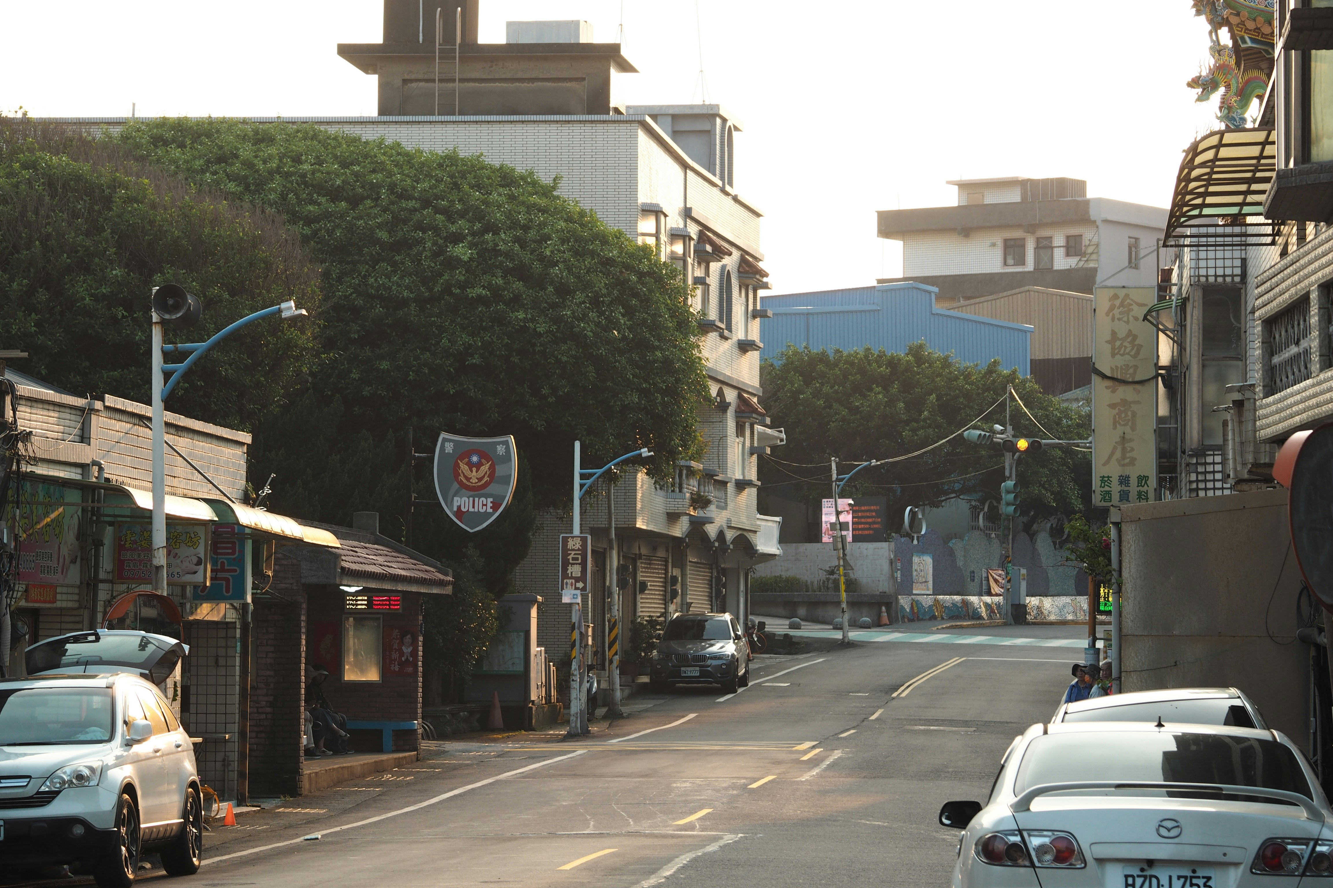 A street with buildings, parked cars, and trees under sunlight.