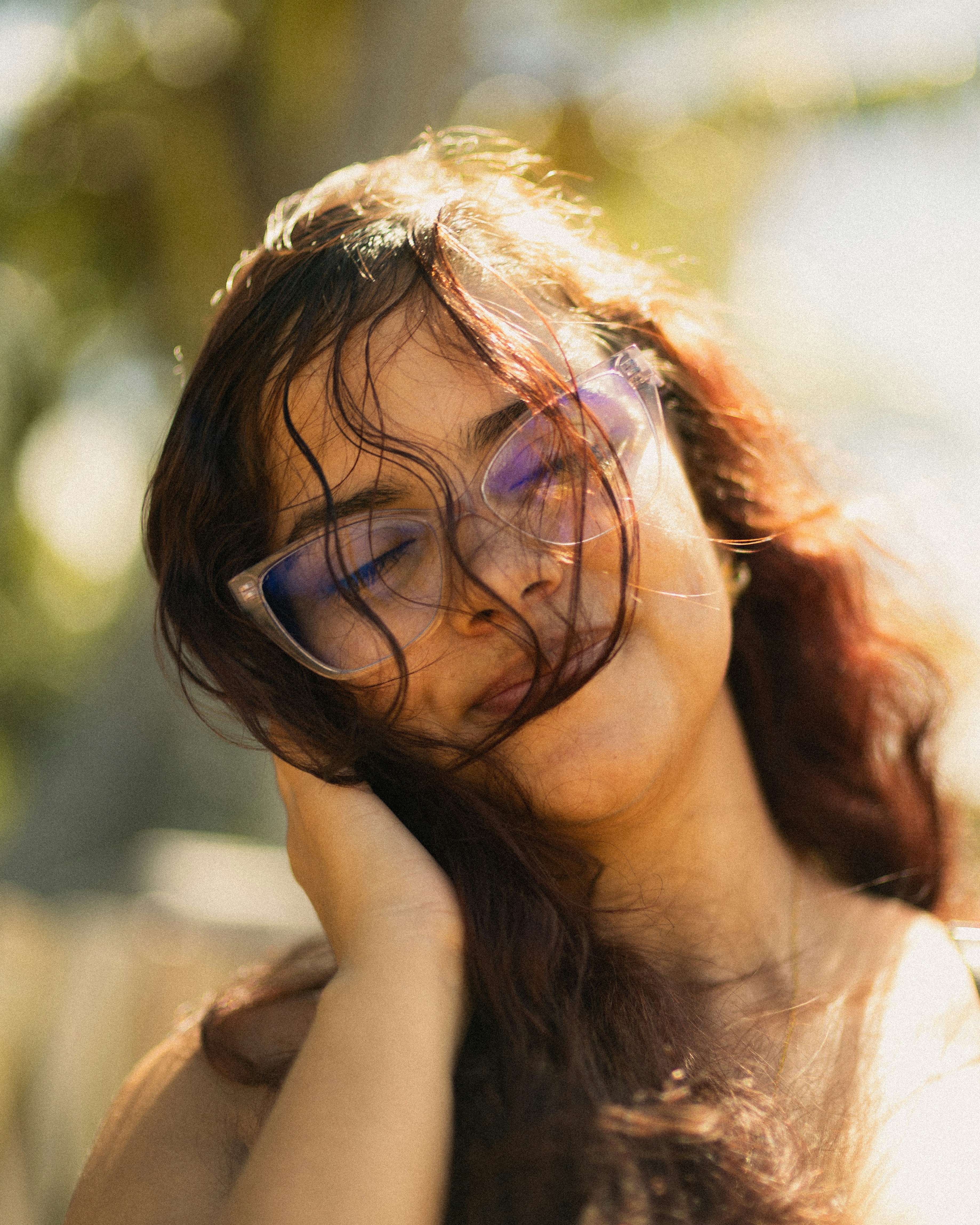 Portrait of a woman with wind-swept hair and tinted glasses, softly backlit with warm bokeh; the image focuses on the face and expression. This is a photographic portrait, not a drawing or painting.