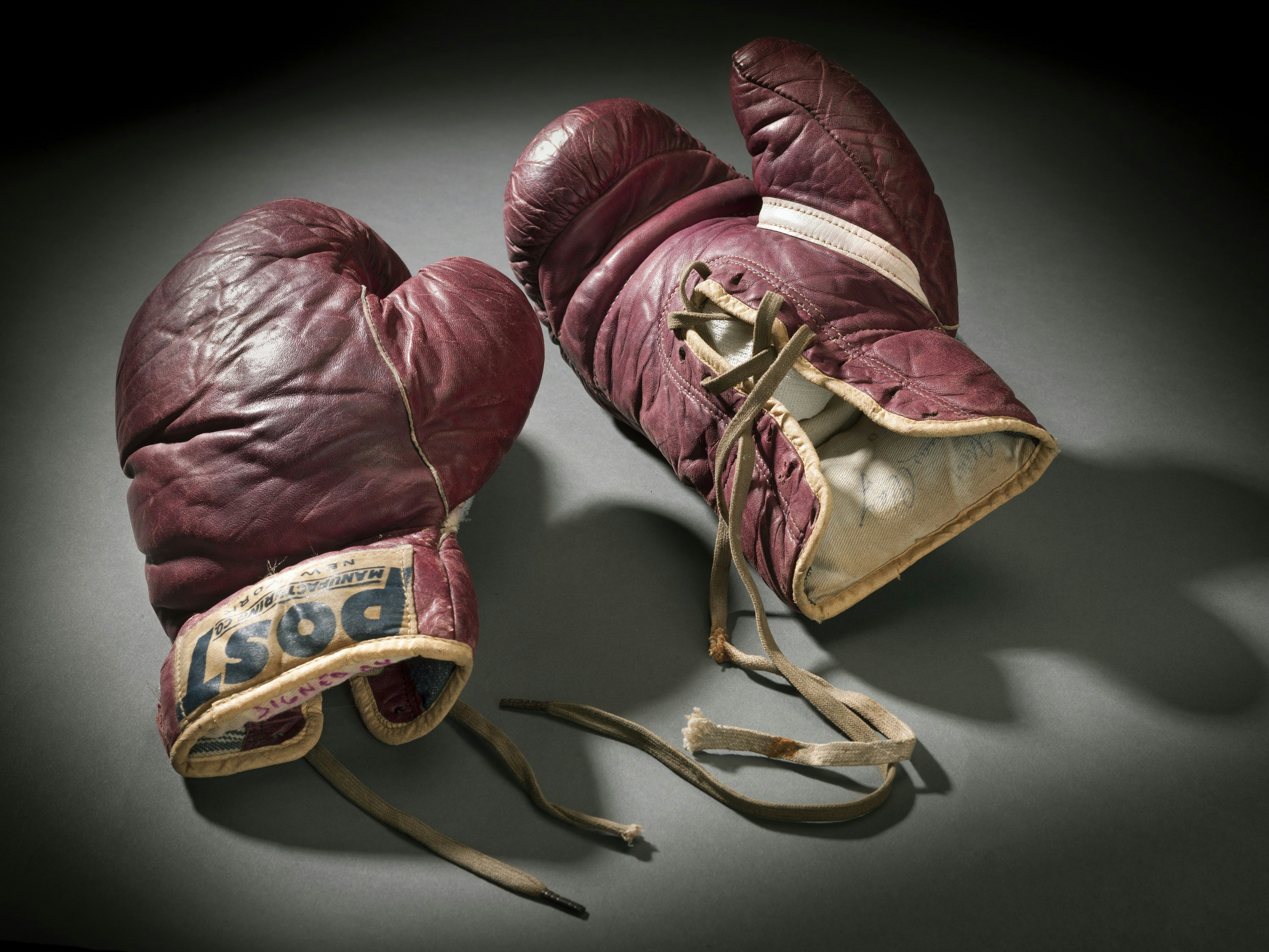 Cassius Clay (Muhammad Ali) Training Gloves, Dark red brown training boxing gloves worn by Cassius Clay (Muhammad Ali) and signed in Louisville, as indicated by the penned note inside the left glove near the wrist. Gloves are edged in faded yellow fabric and tied with dark, faded strings. Stitching throughout the glove is in white thread. On the outside fronts of both gloves below the wrist are manufacturer's labels. Post Manufacturing Co., circa 1960, Record ID nmaahc_2012.173.3ab