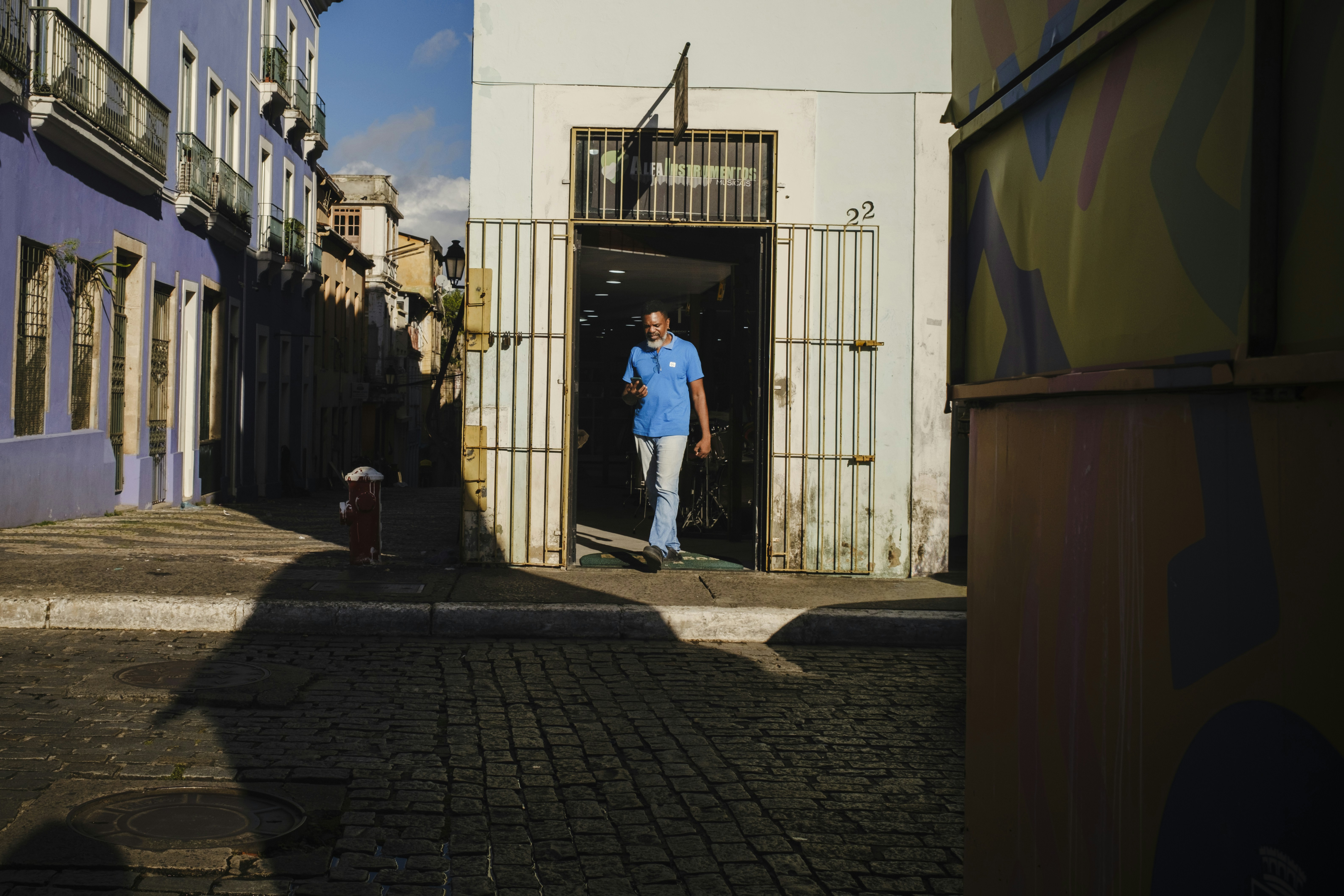 A lone traveler emerges from a weathered gate into sunlit cobbles