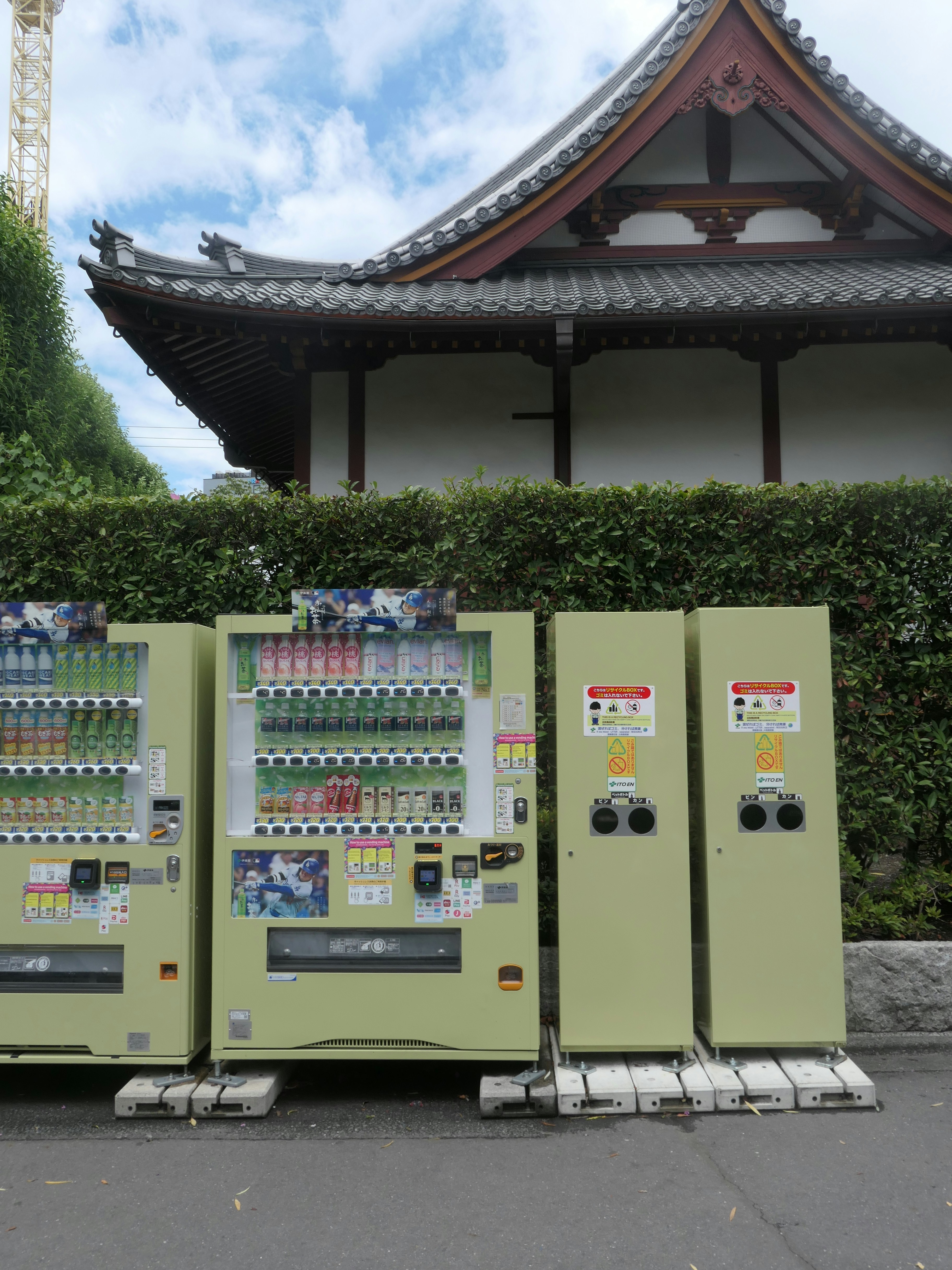 Row of pale-green vending machines stands before a traditional tiled roof and trimmed hedge, capturing a quiet urban moment.