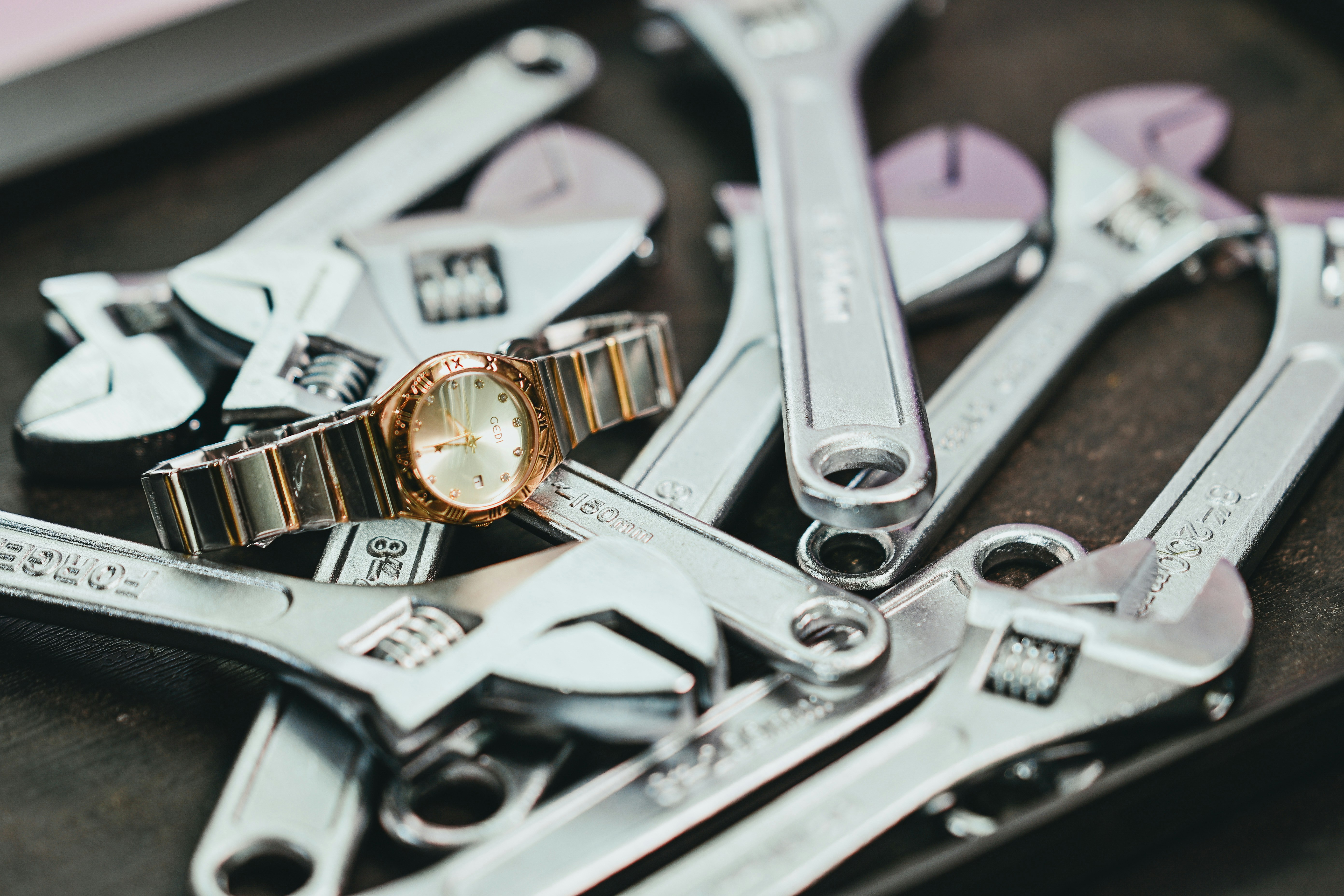 A gold-toned wristwatch sits among metallic wrenches and spanners, resting on a dark surface; the scene emphasizes craftsmanship and tool-filled surroundings.