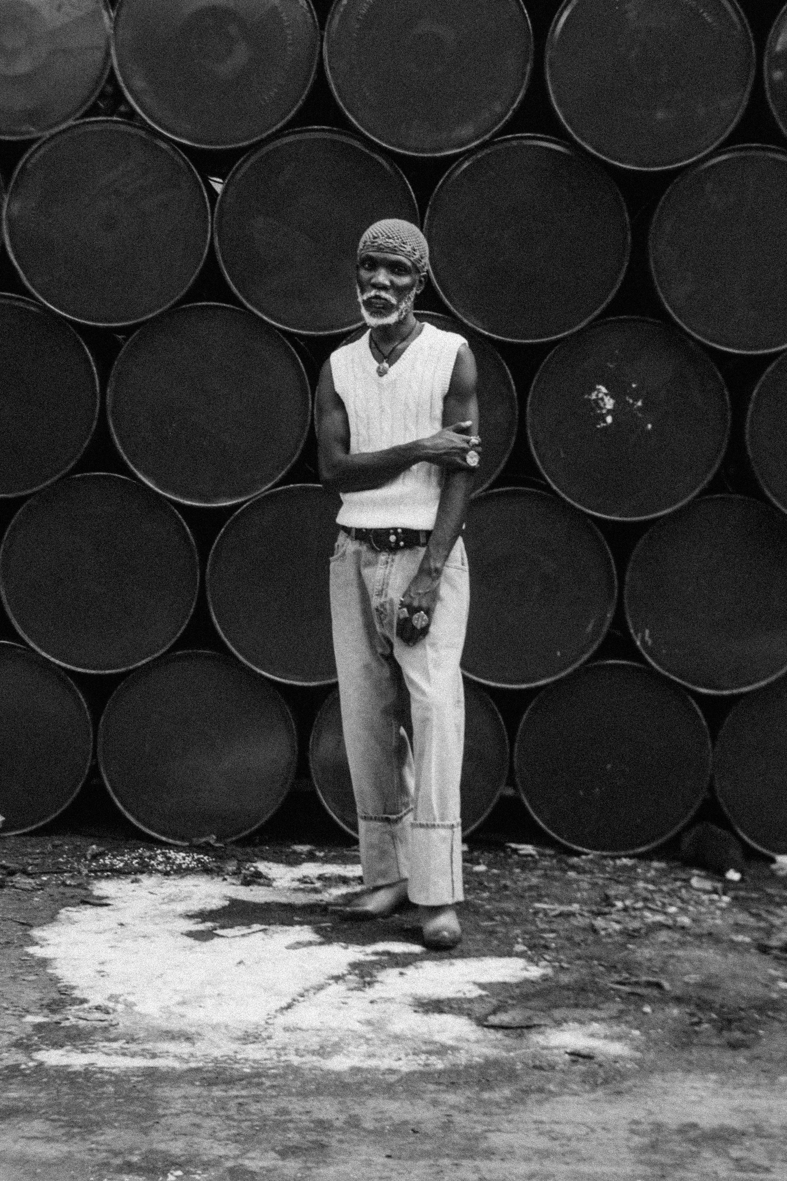 Man in white sleeveless shirt stands before stacked industrial drums