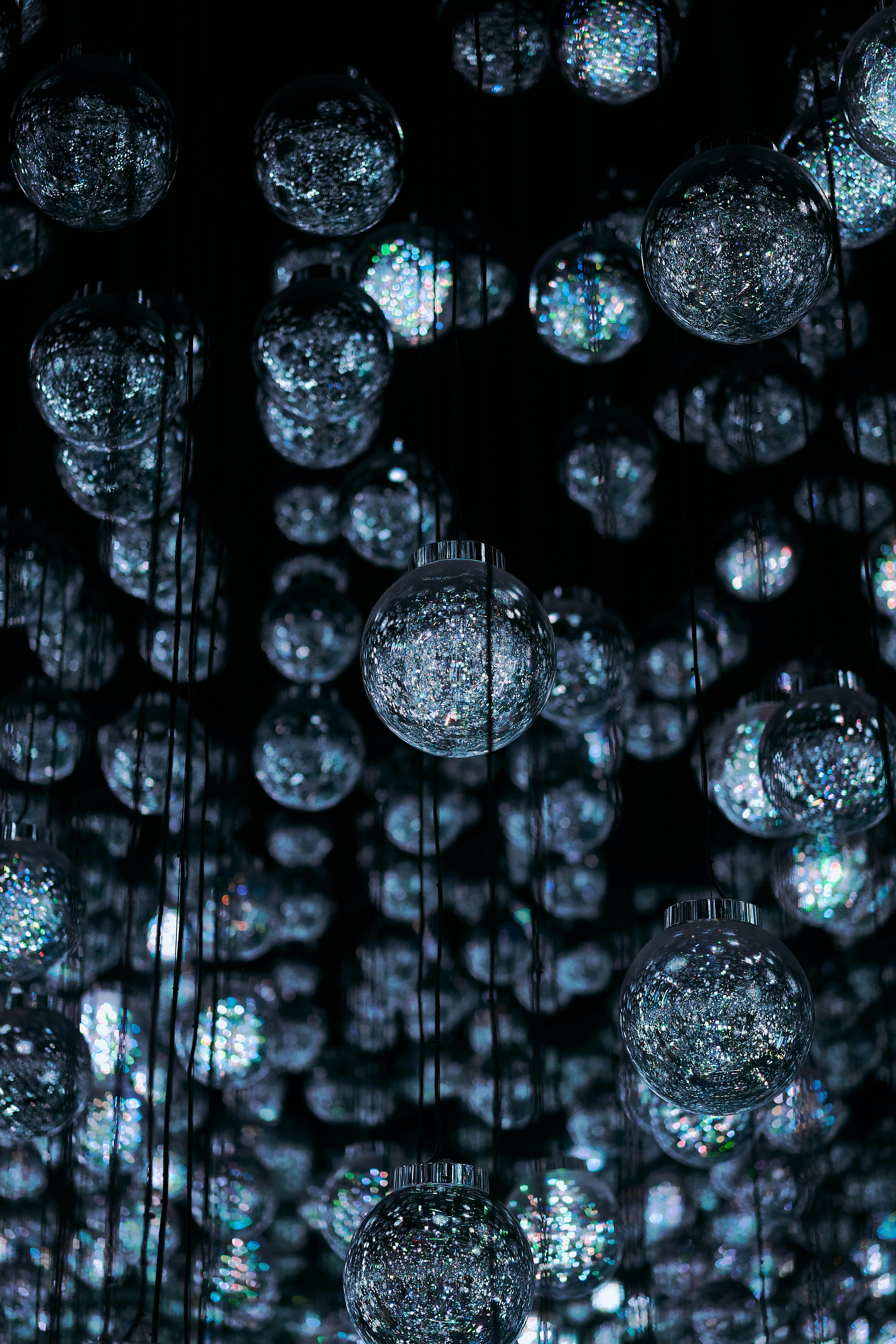 A dark installation of numerous glass orbs suspended by thin cords, each filled with glittery specks that reflect subtle blue light. The composition emphasizes depth and repetition, creating a floating constellation effect.