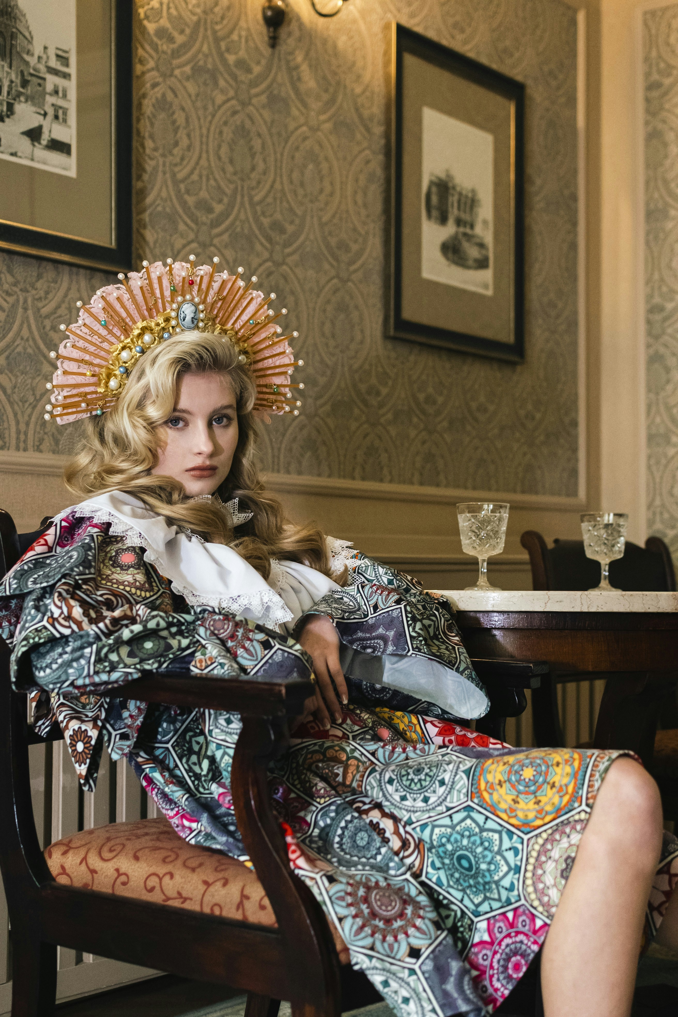 Elegant portrait of a woman in a richly patterned robe and an ornate headdress seated in a vintage interior. A framed wall and glassware on a table suggest a refined, historical setting.
