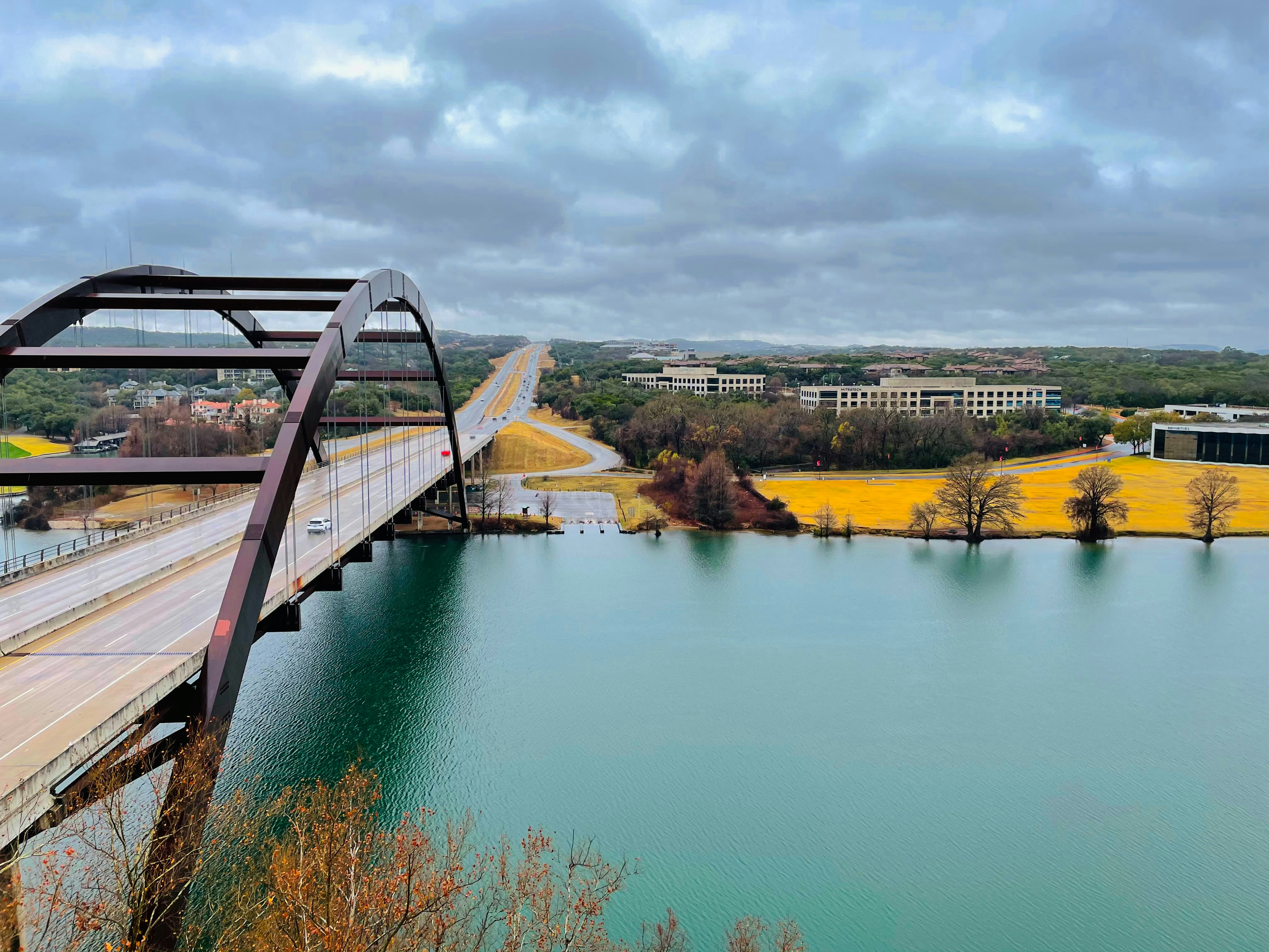 Bridge spans a calm river with a bright yellow meadow beyond and a cloudy sky above, capturing a modern arc structure against a tranquil landscape.