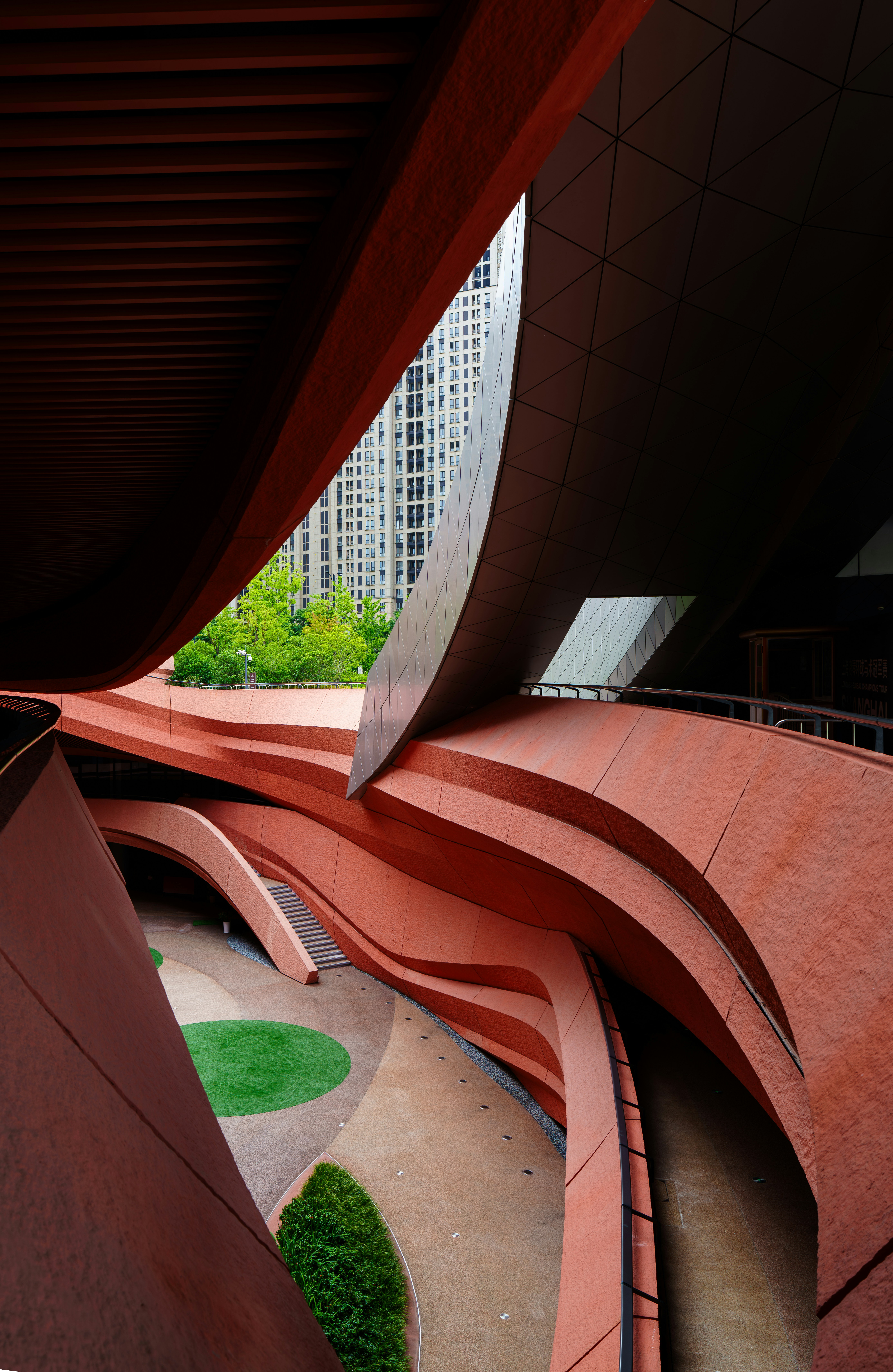 Sinuous red architectural ramps carve through a modern structure, with green courtyard glimpses and distant buildings beyond. This image captures an abstract urban landscape with bold curves and contrasting textures.