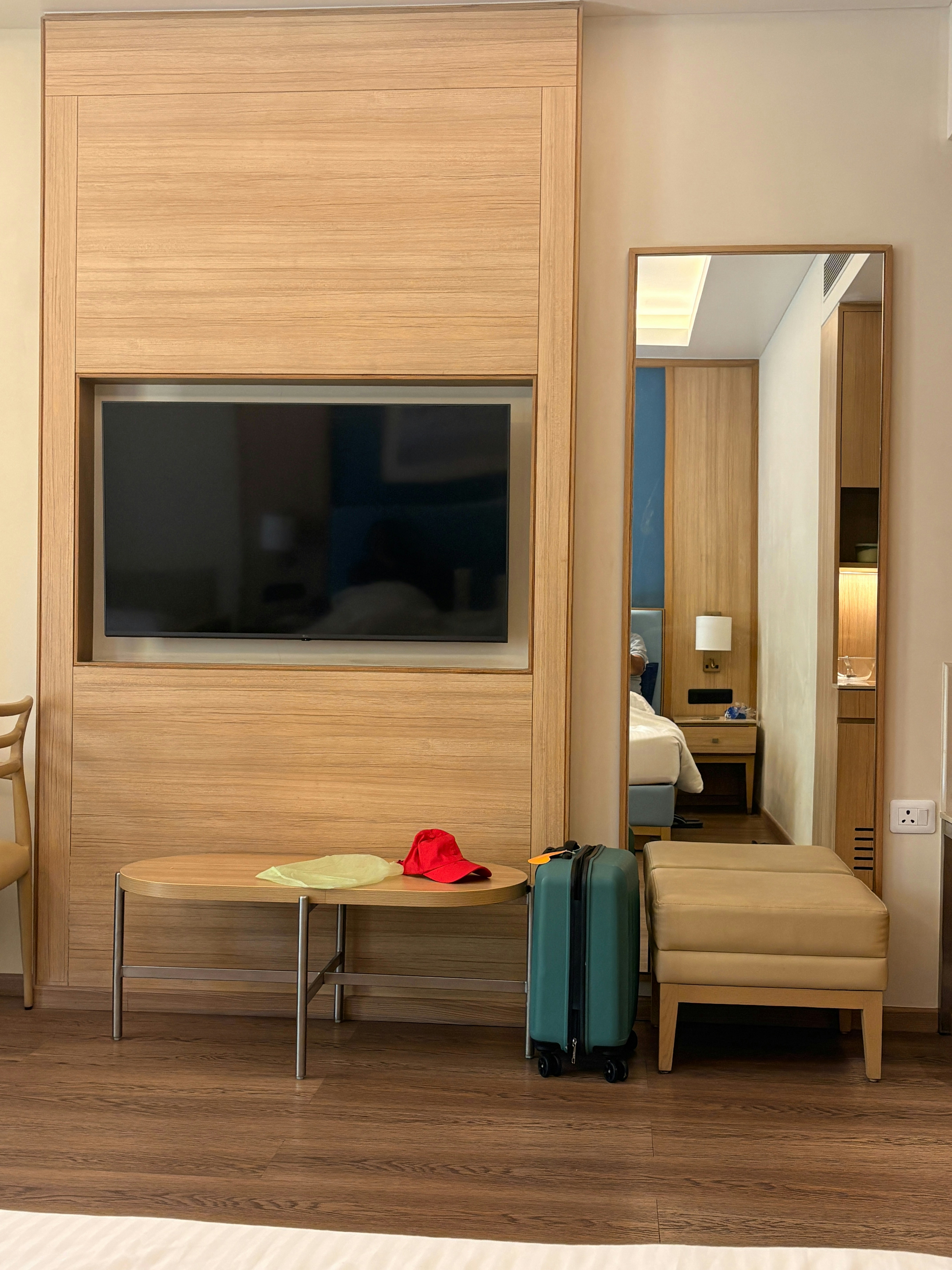 Modern hotel room with tv, mirror, suitcase, and wooden furniture.