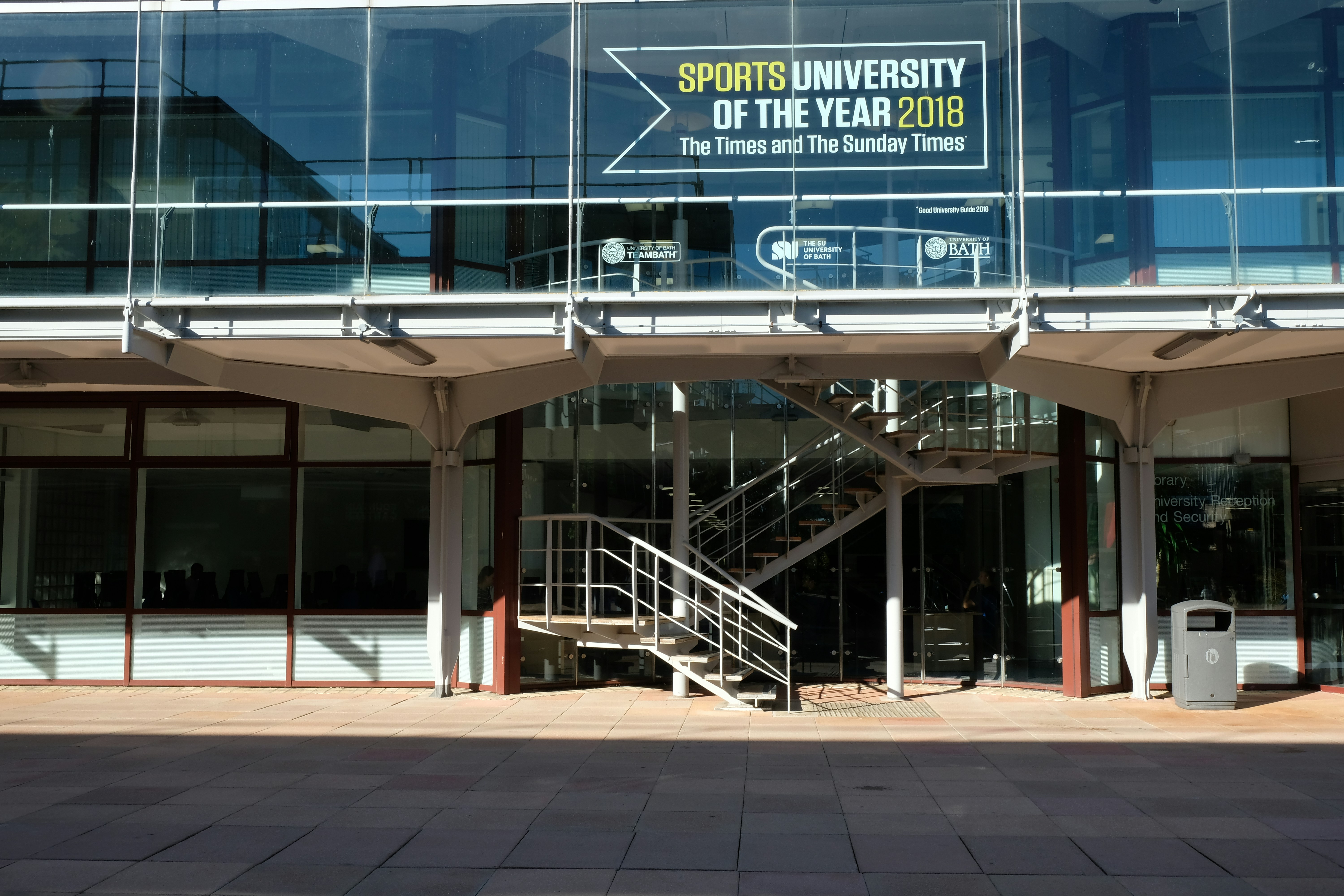 Sunlit glass-front building with a central staircase and banner above, showcasing a modern sports facility entrance.
