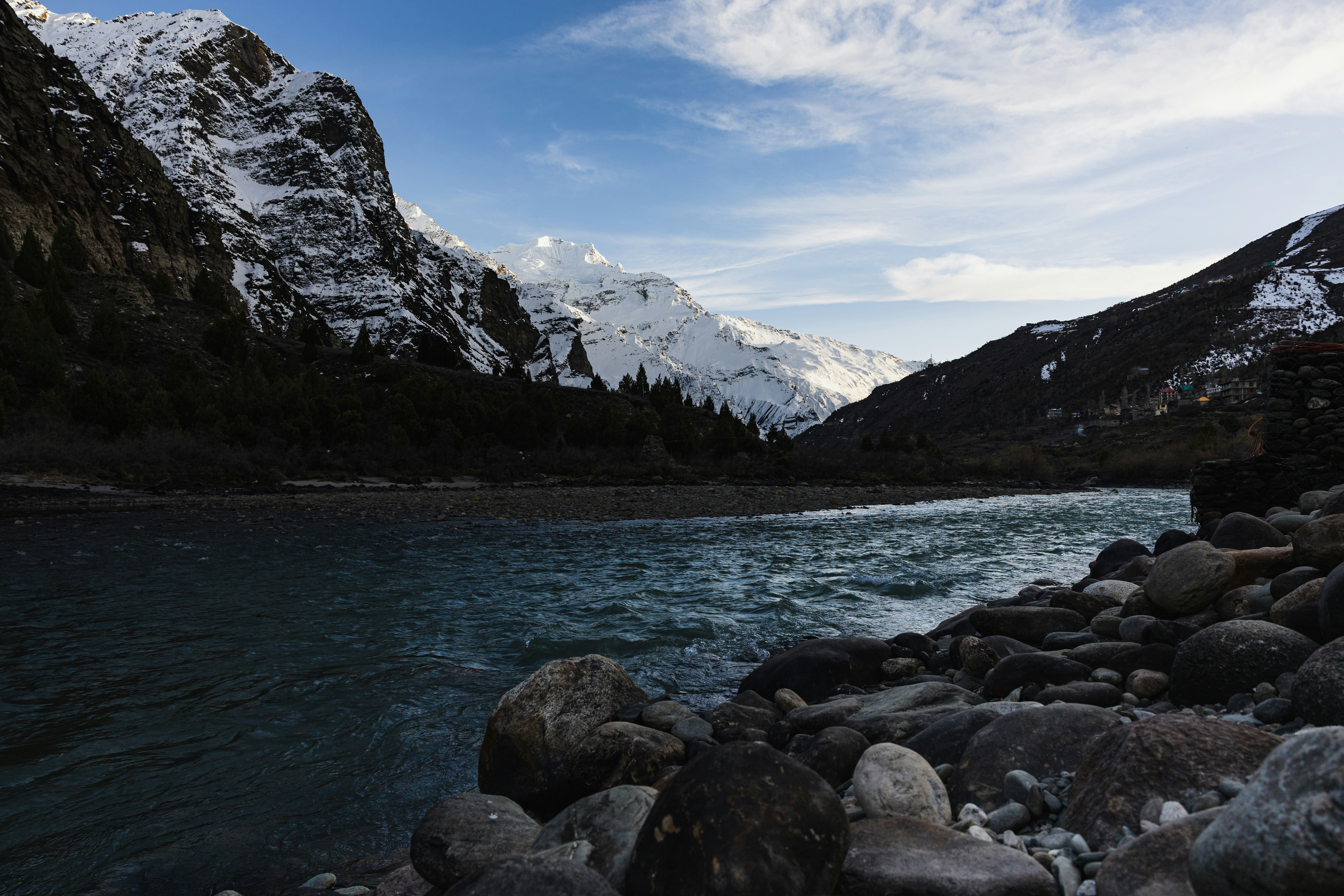 Icy river winds through a rocky shore with dark pines at the base of towering snow-covered cliffs under a clear blue sky.