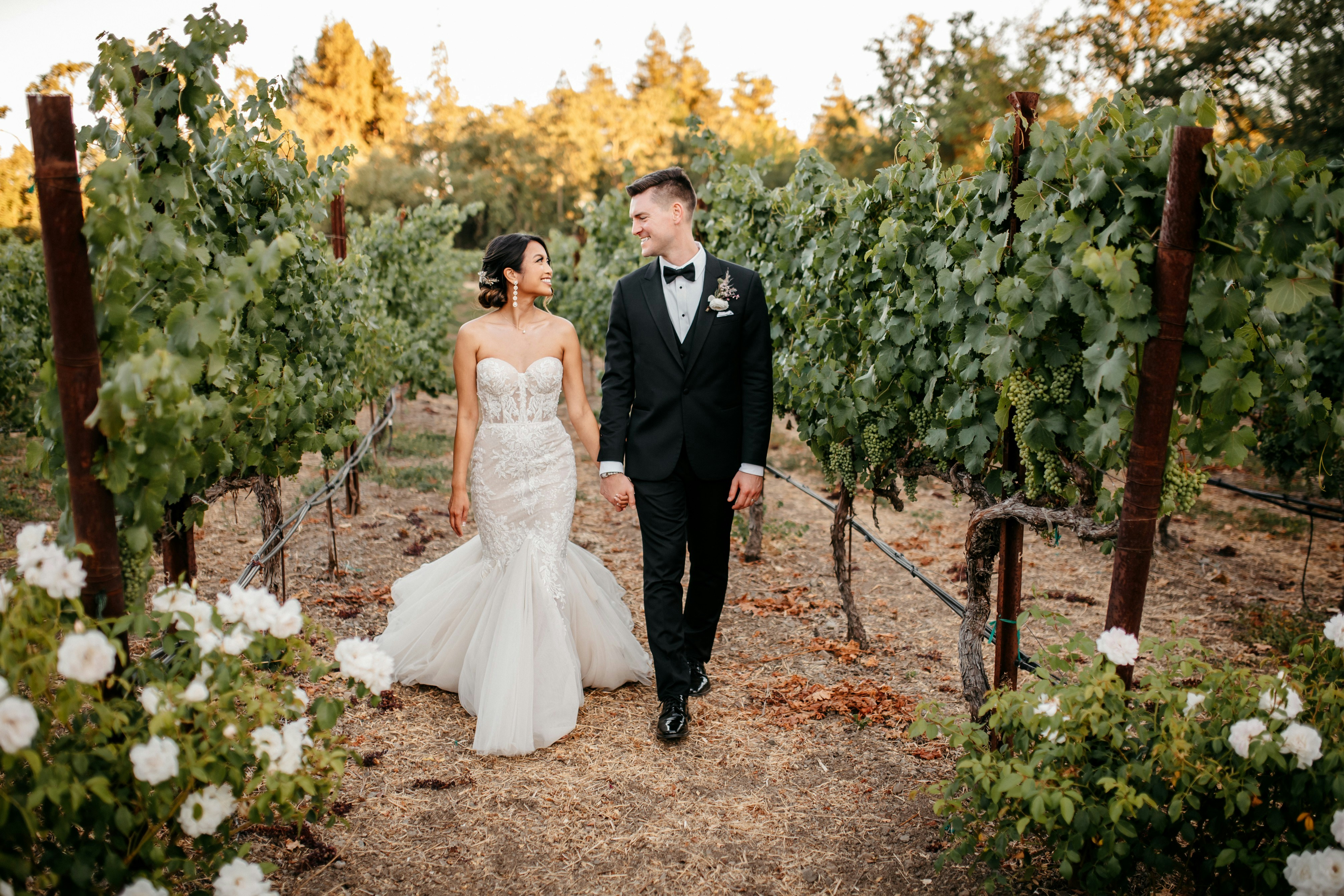Newlyweds stroll through a sunny vineyard with white flowers.