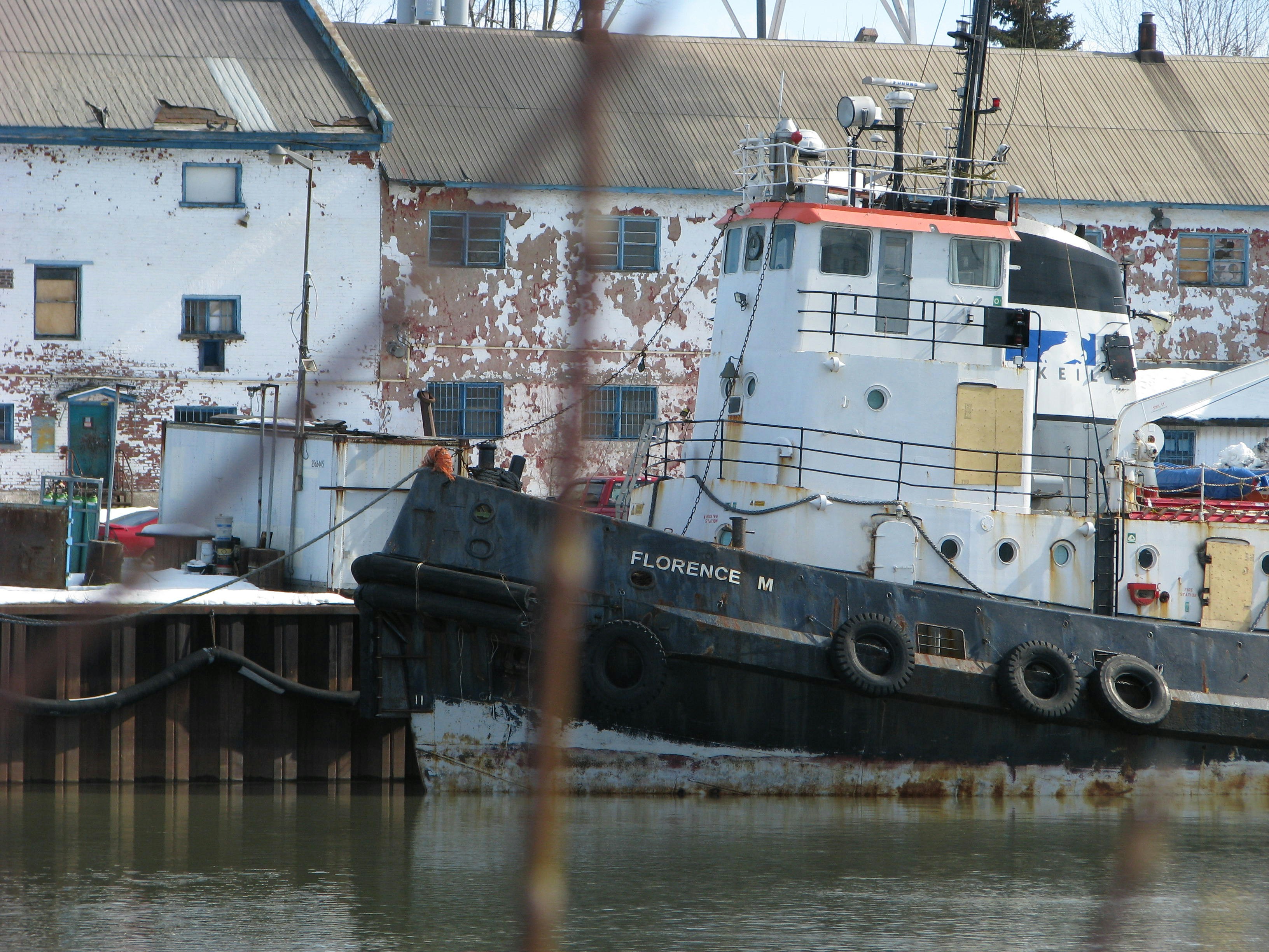 A weathered tugboat named 'FLORENCE M' sits alongside a gritty, rust-streaked dockyard building with peeling white paint. The scene emphasizes the vessel’s dark hull, foredeck details, and calm water reflection.