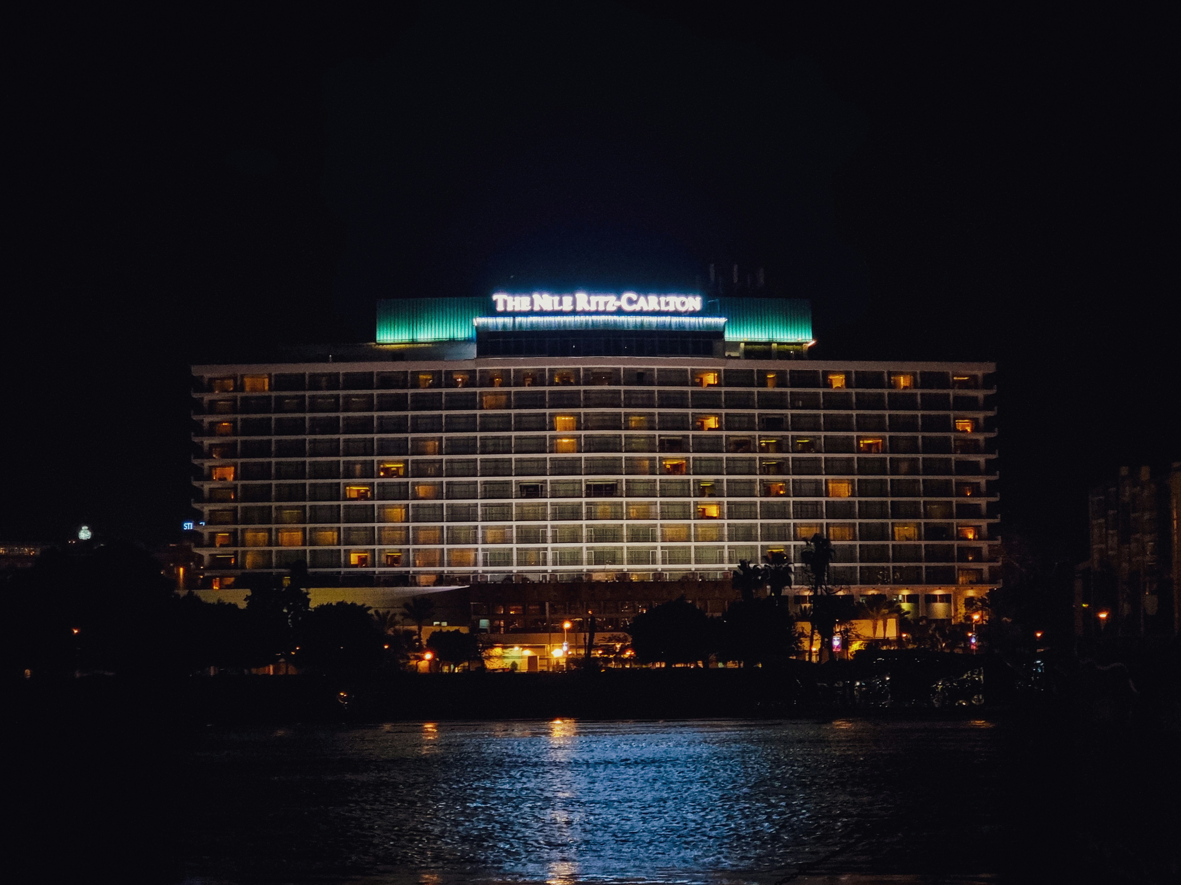A glowing architectural landmark stands out against the dark city night, with its grid of illuminated windows and neon sign reflecting on the calm waters below.