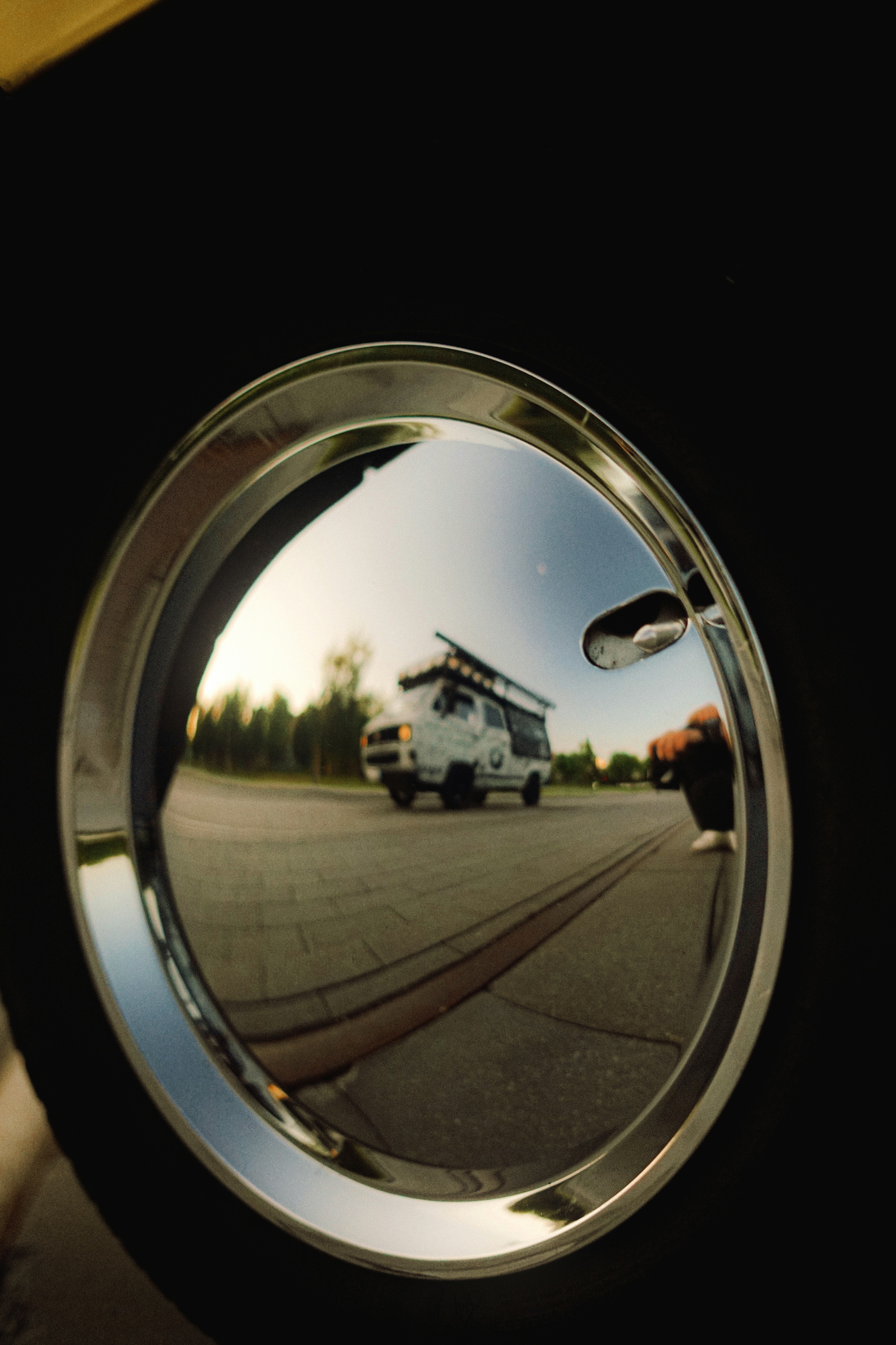 A close-up view through a chrome circular reflector reveals a parked van with a rooftop rack and a person nearby, set on a paved area with greenery in the background. The image is a chrome reflection shot.