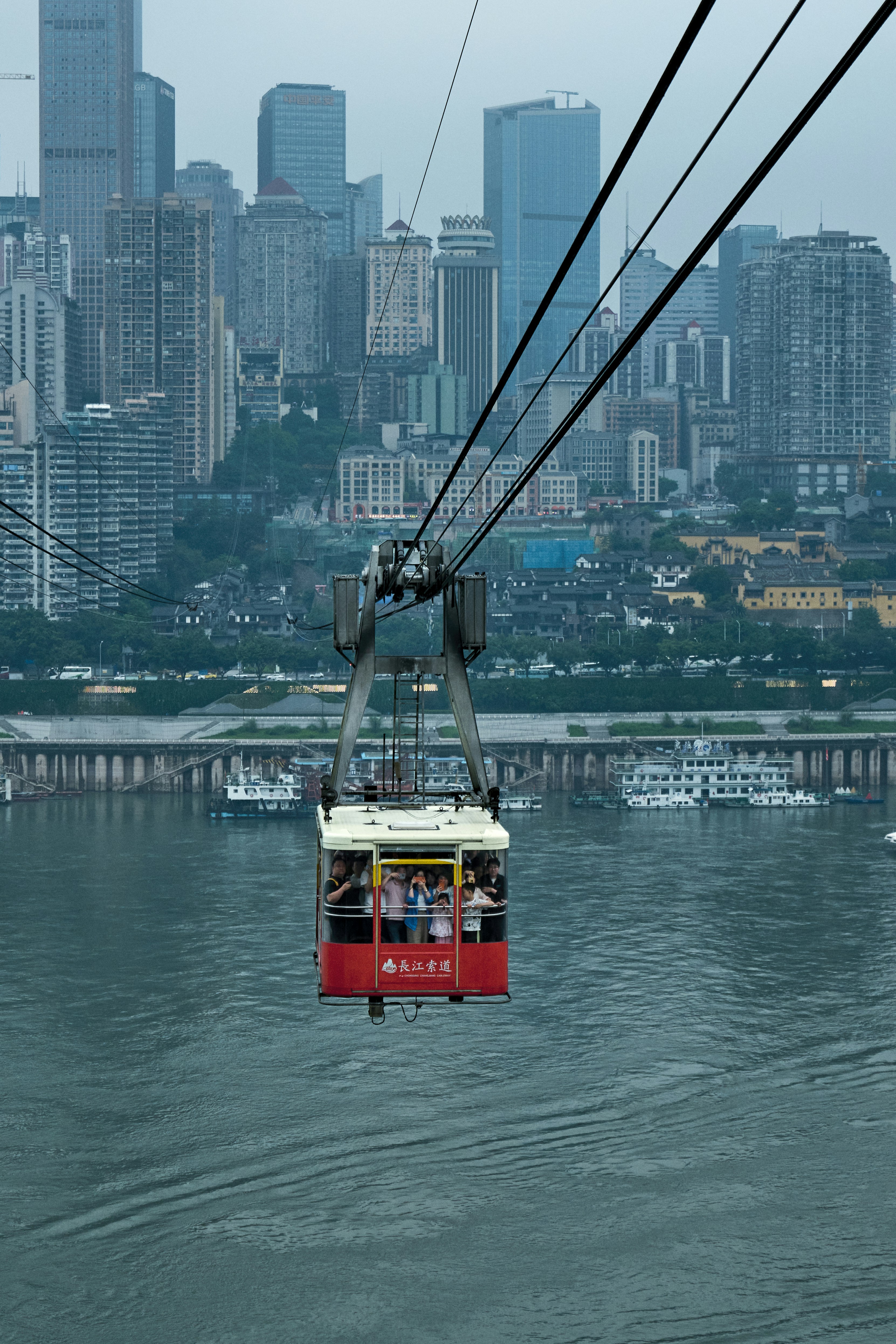 Red cable car crosses river, towering city skyline in background.