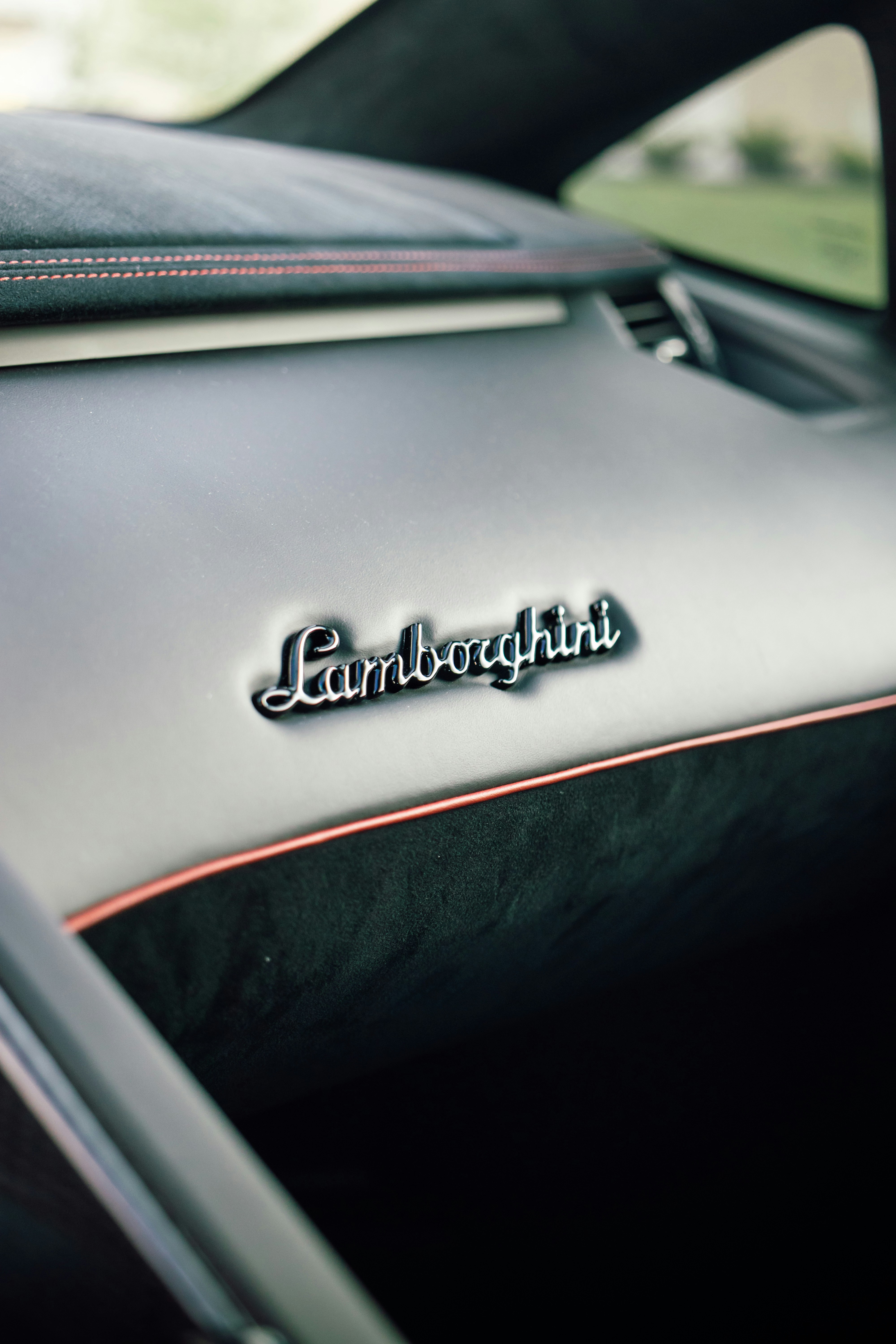Close-up of a Lamborghini emblem on the dashboard, with soft background blur and red stitching accents inside a car cockpit.