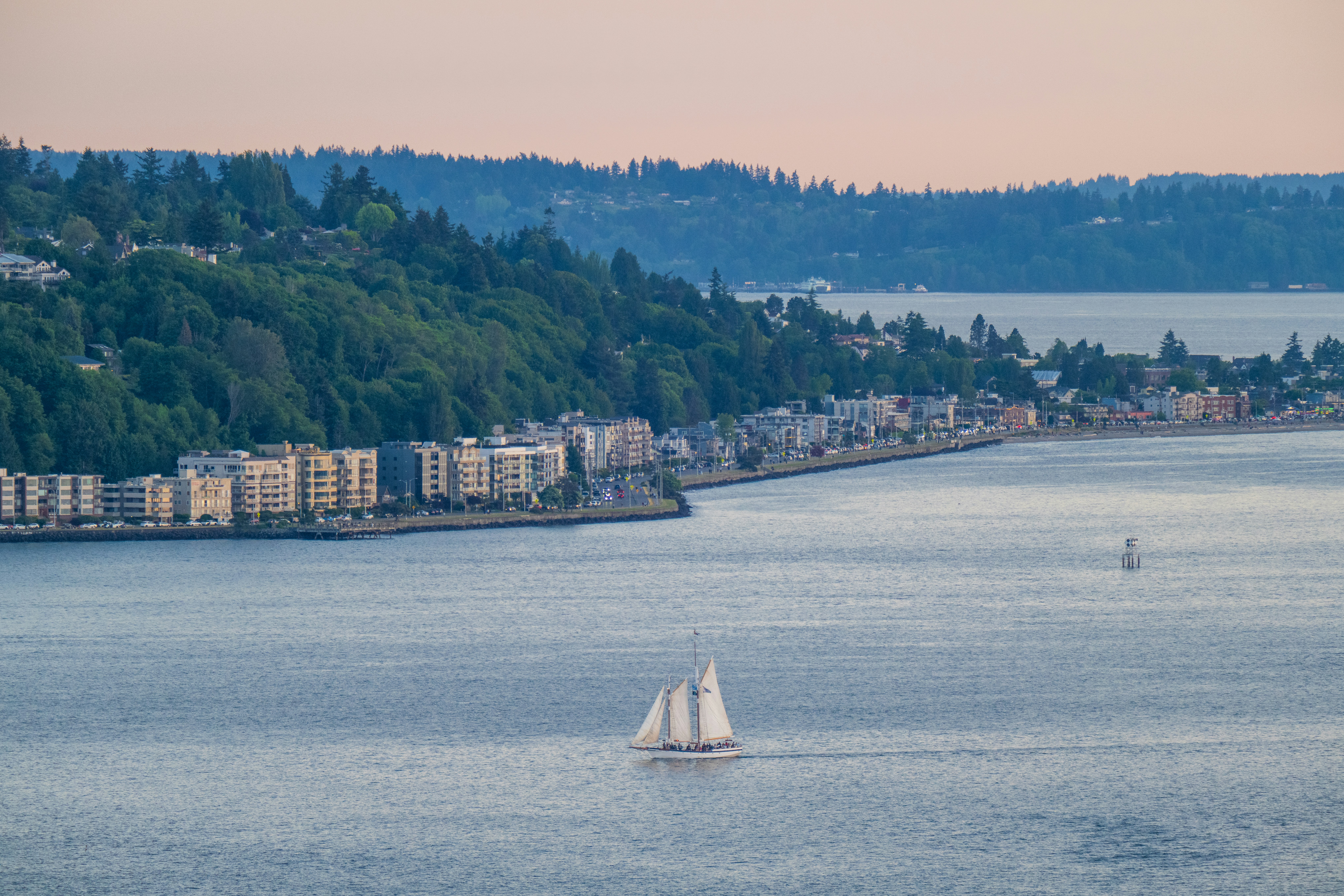 Sailboat sailing past a coastal town and tree-covered hills.