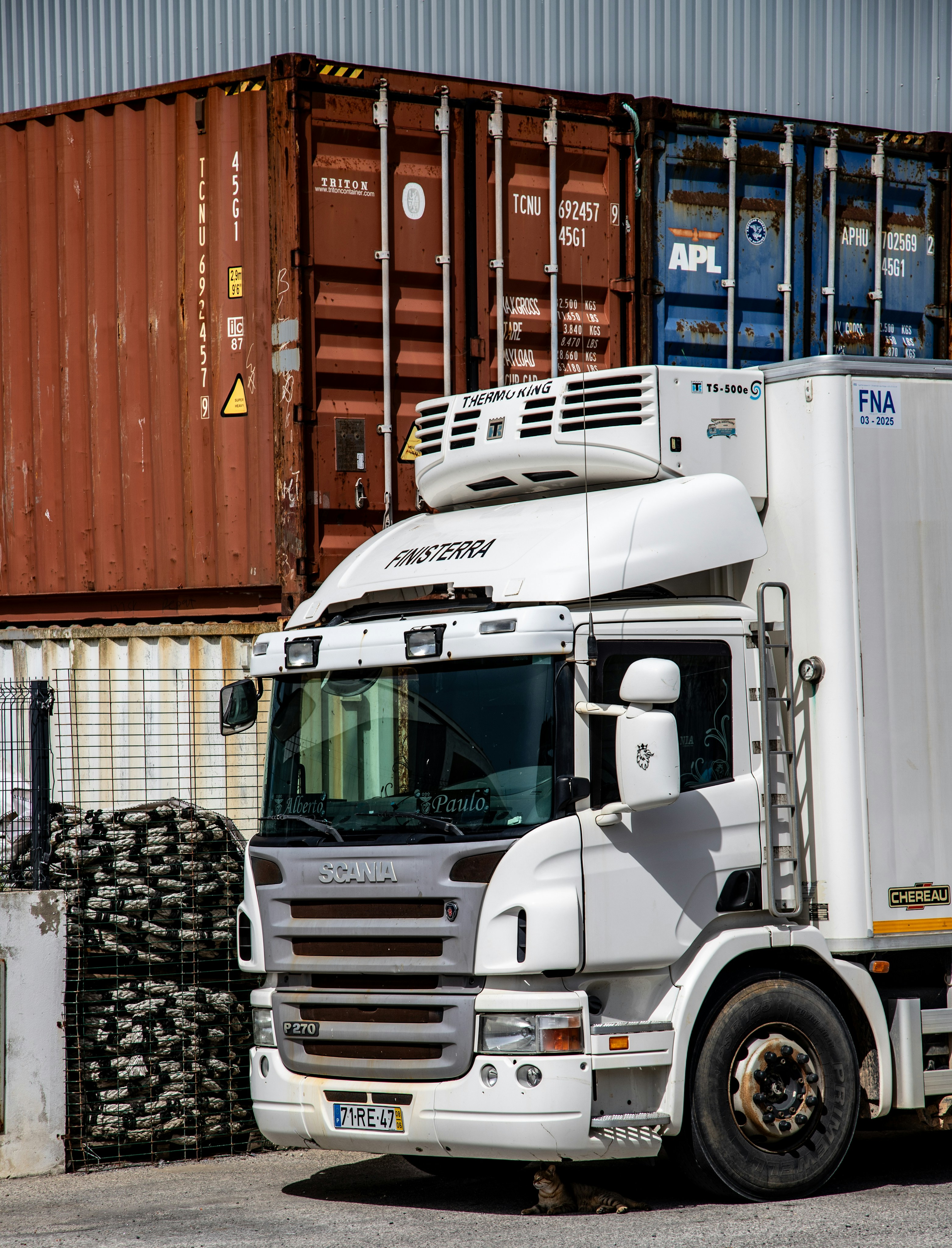 A white tractor-trailer rests beside stacked cargo containers under a sunlit yard