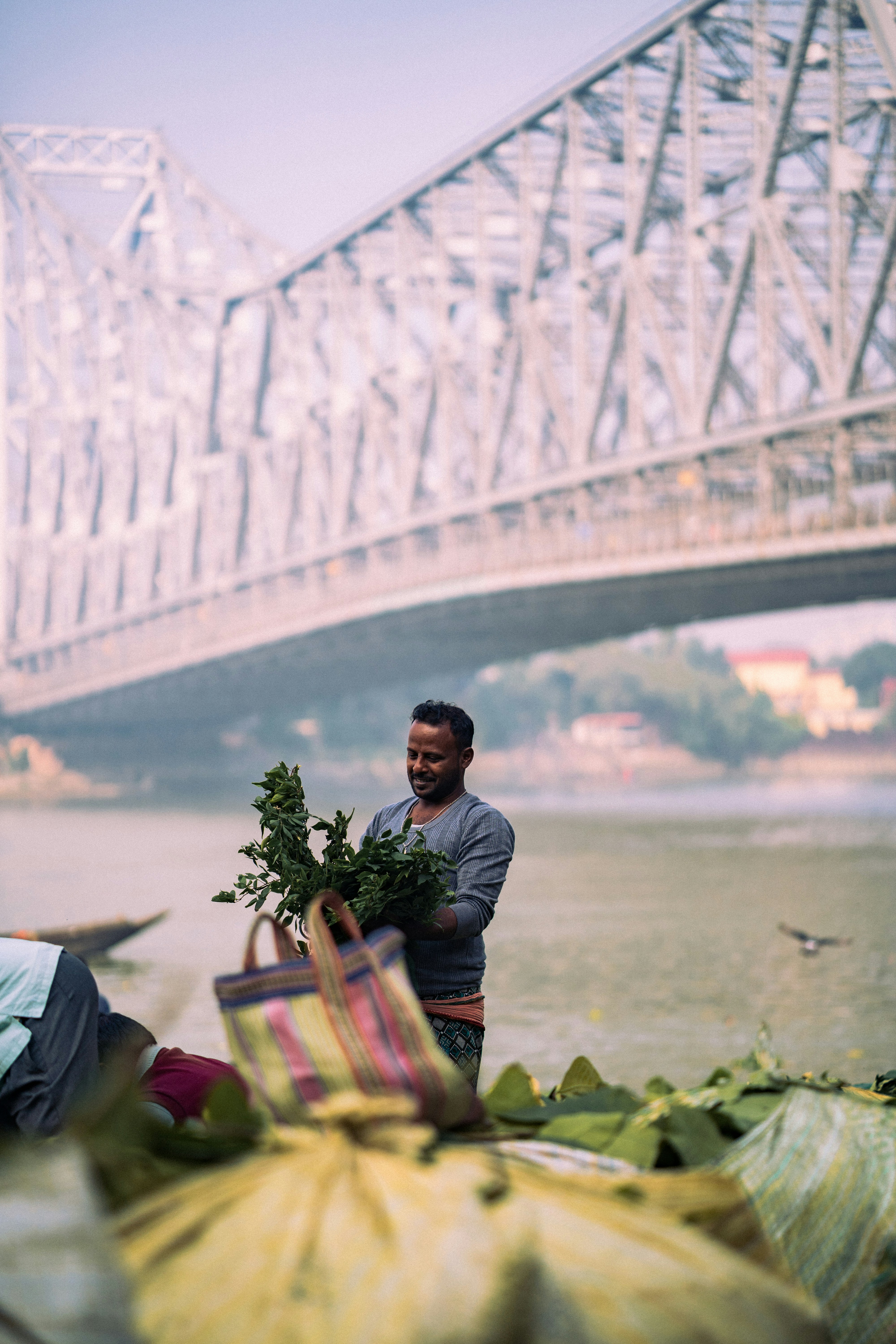 Vendor with leafy greens by a river under a steel arch bridge