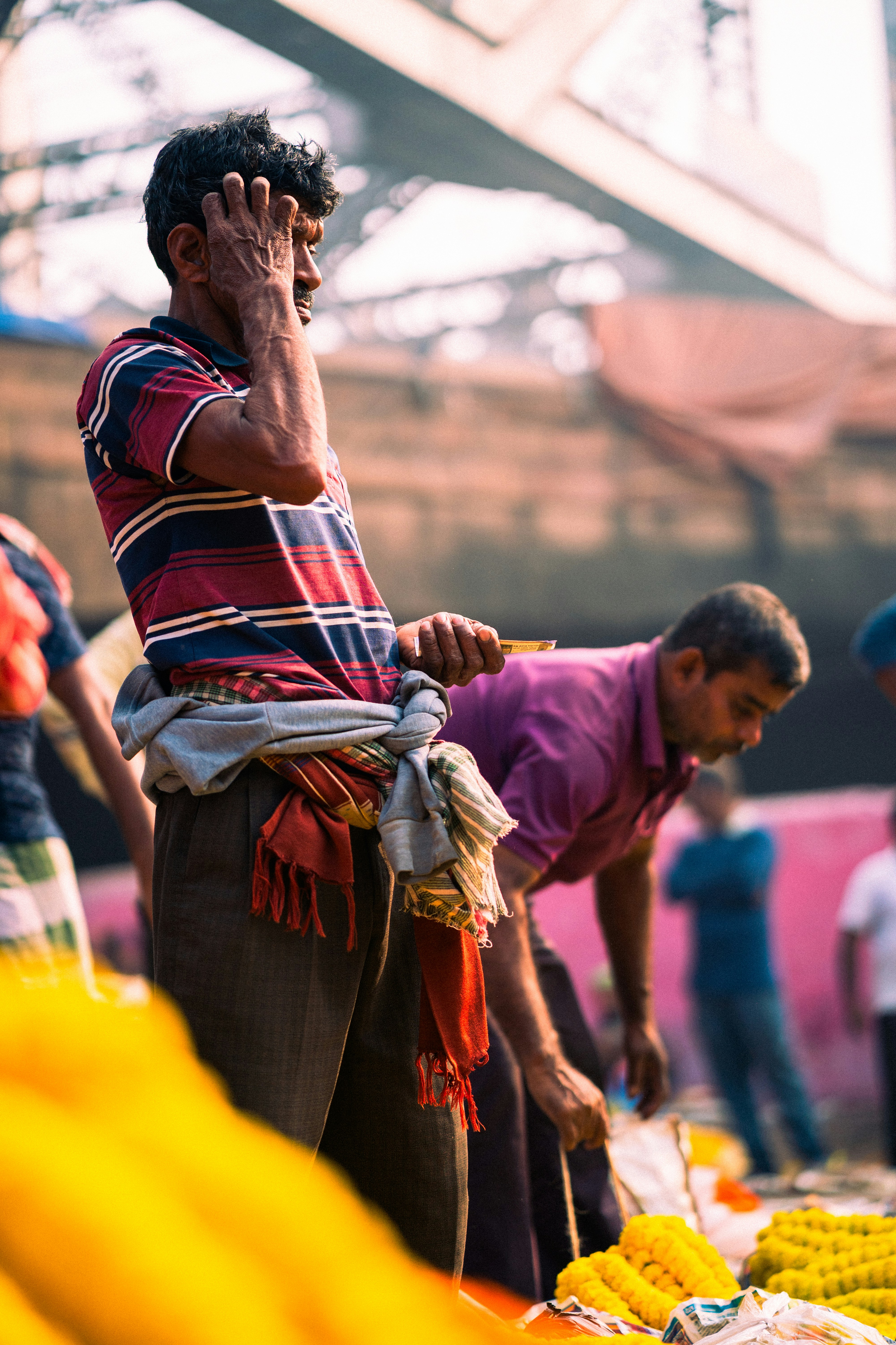 Man in striped shirt pauses amid a flower stall under a distant steel bridge