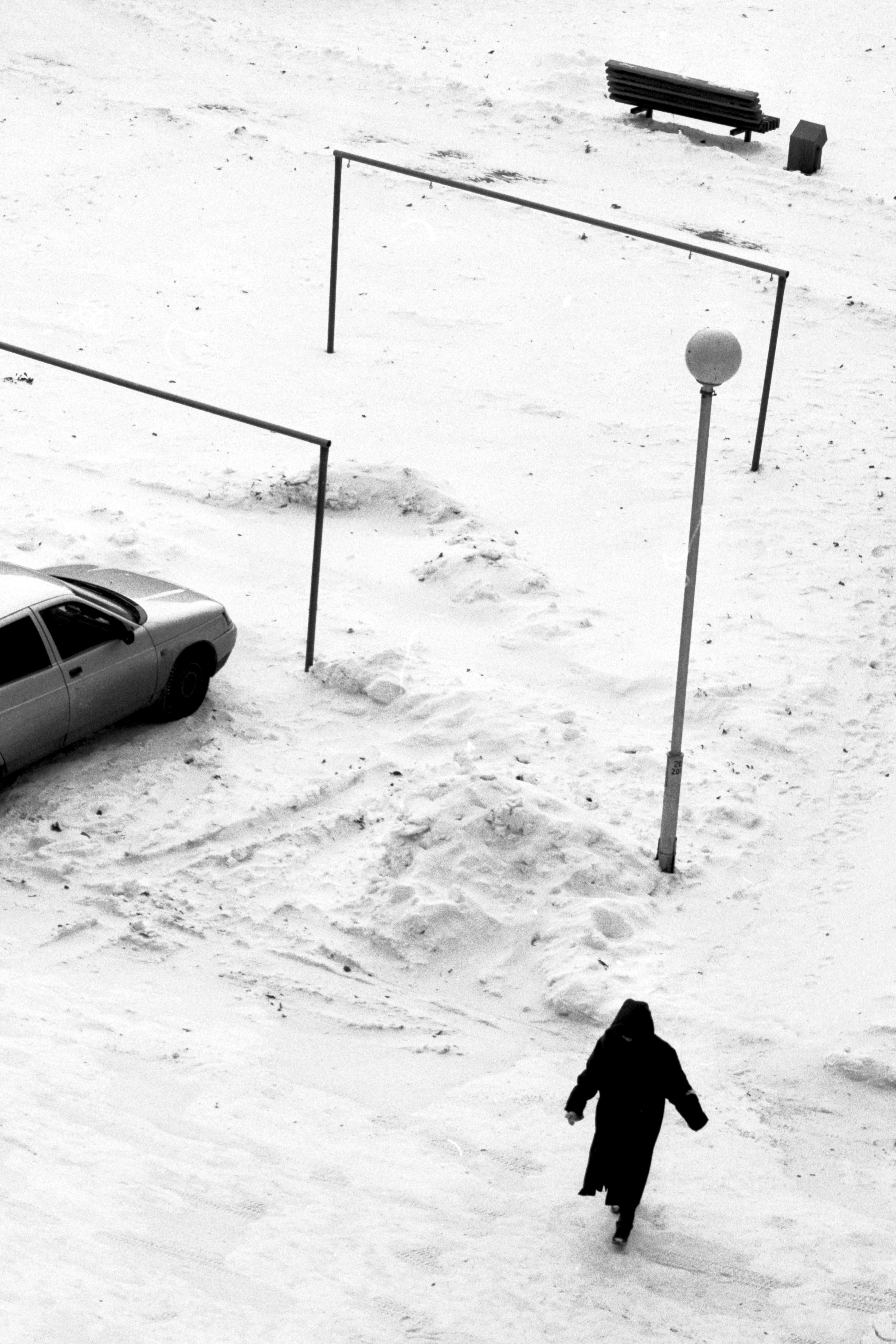 Person walks in snow past car, lamppost, and bench.