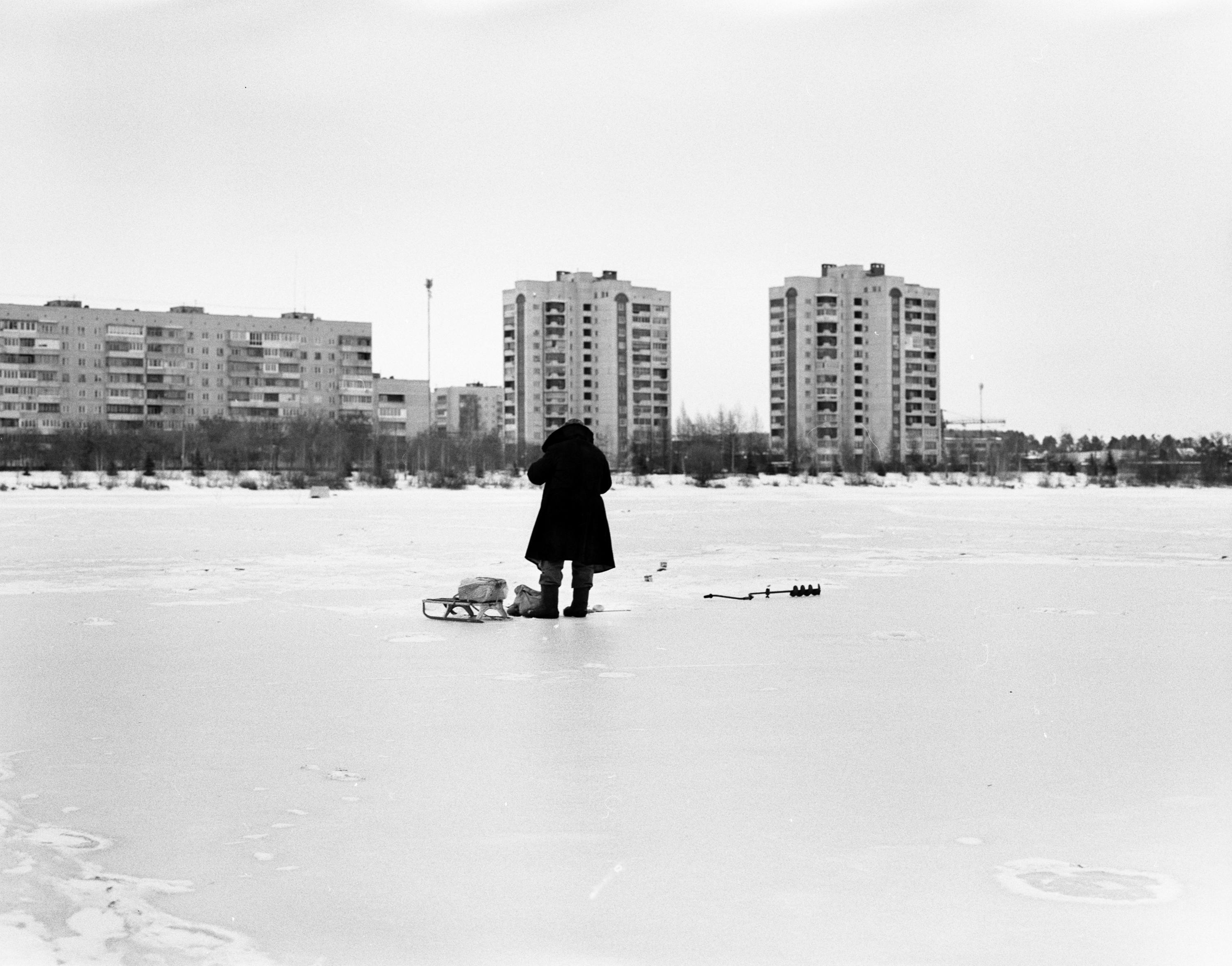Ice fisher on a frozen lake with distant city buildings.