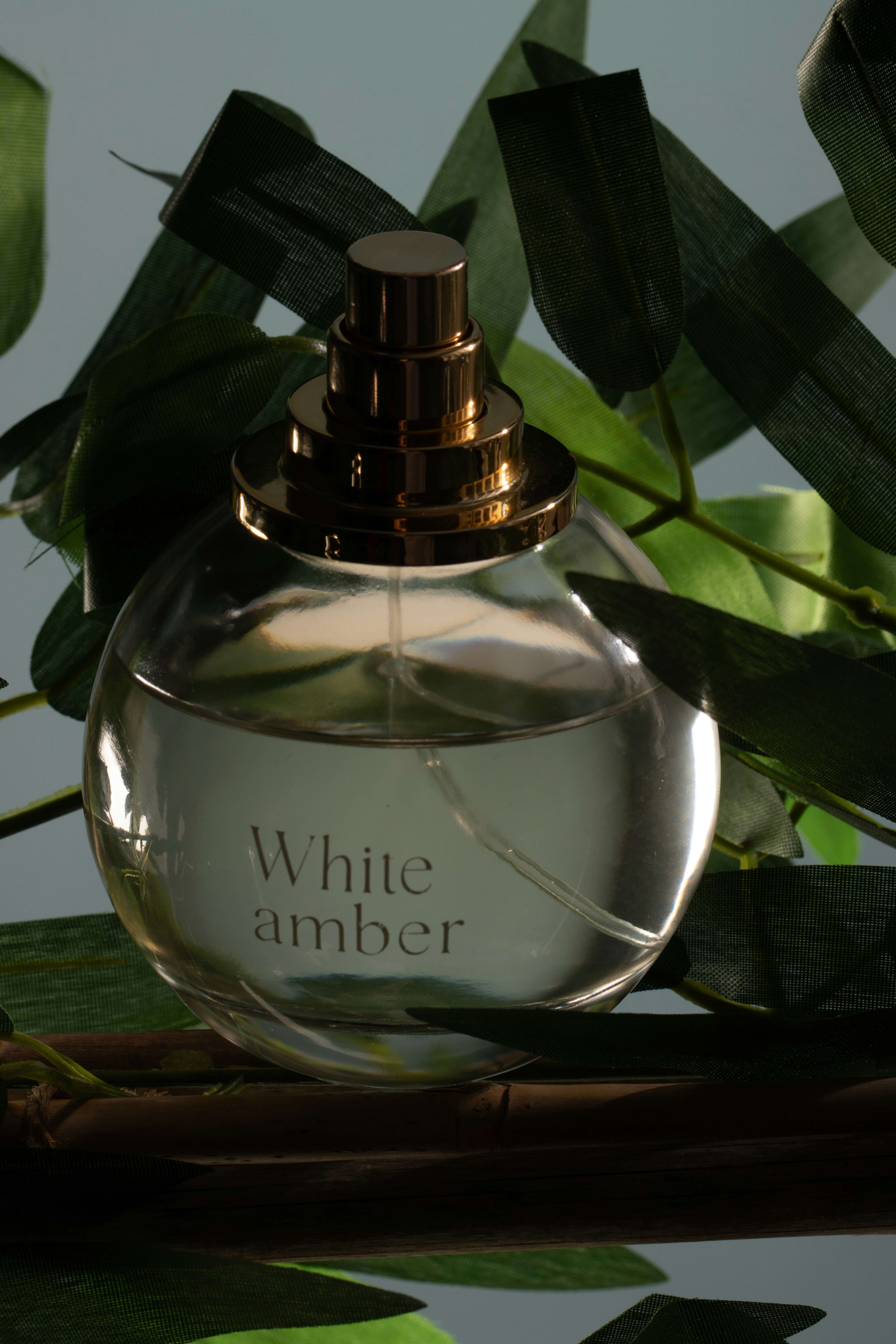 Square-shaped perfume bottle with metallic cap sits among dark-green foliage on a wooden stem, the translucent liquid visible through the glass. A logo-like label reads 'White amber' in soft typography.