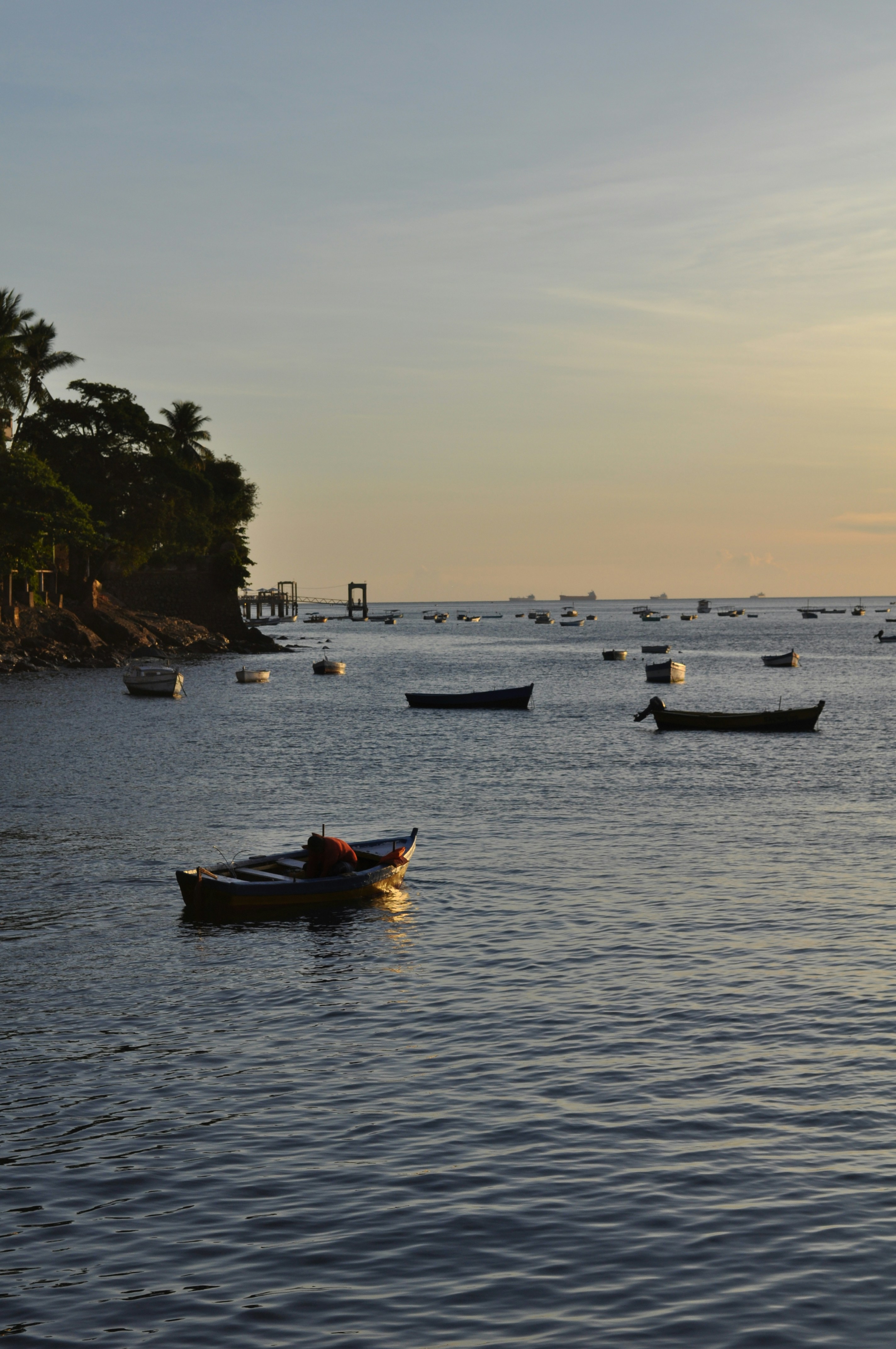 Sunlit harbor with several small boats drifting on calm water beside a rocky, tree-lined shore at sunset.