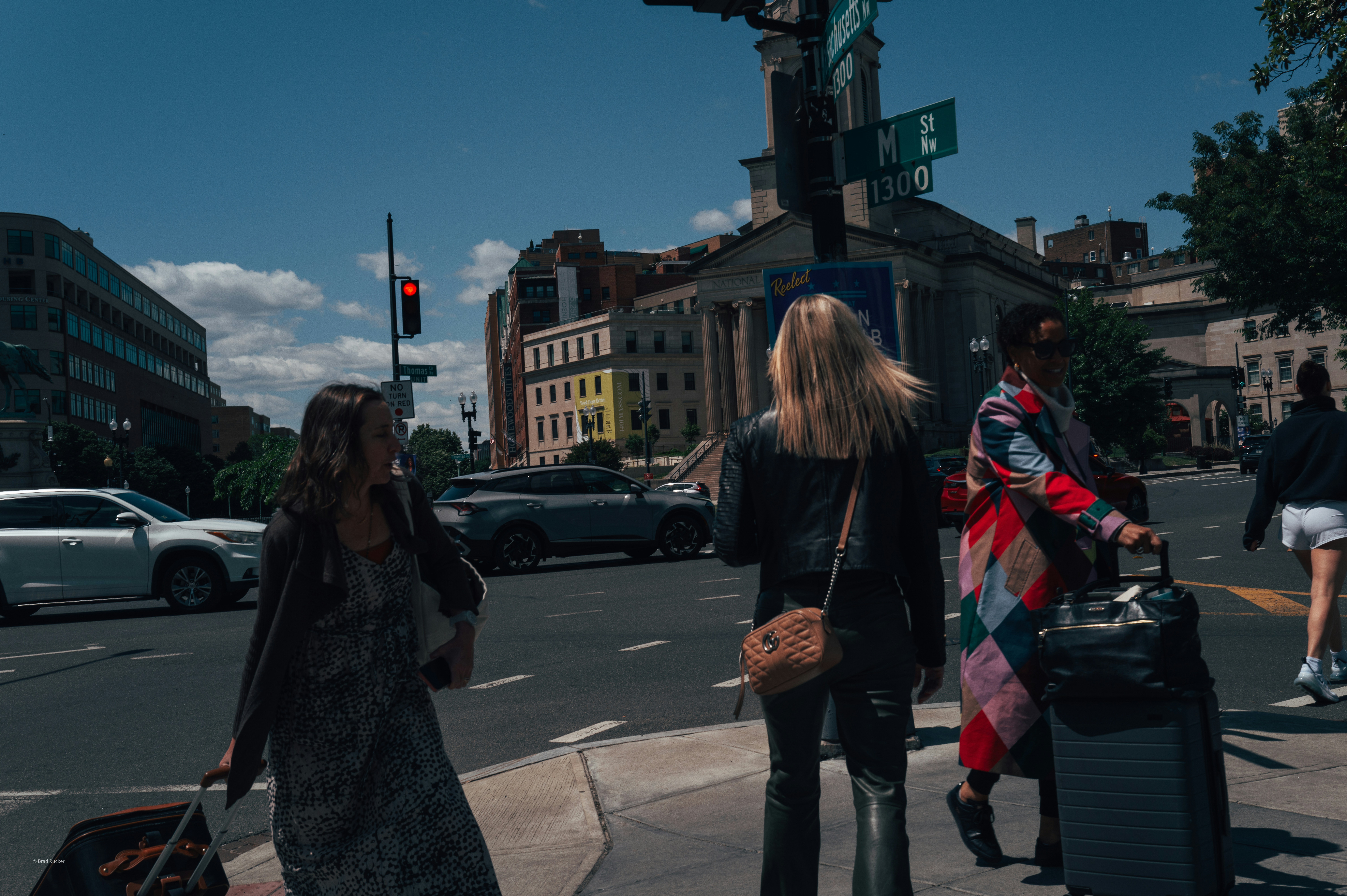 Three women with luggage crossing a sunny city street.