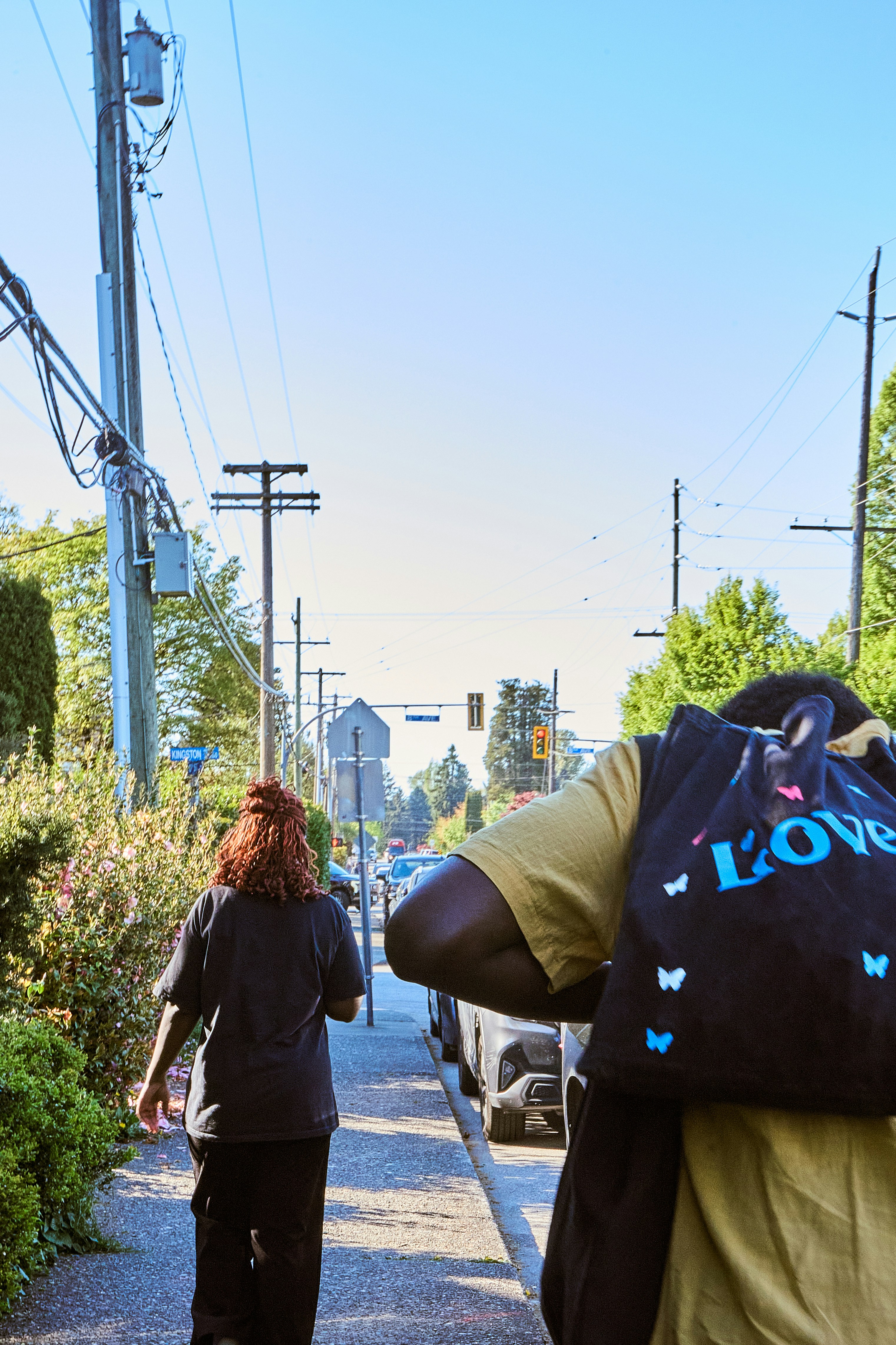 Two pedestrians on a sunlit sidewalk with a backpacked figure in foreground
