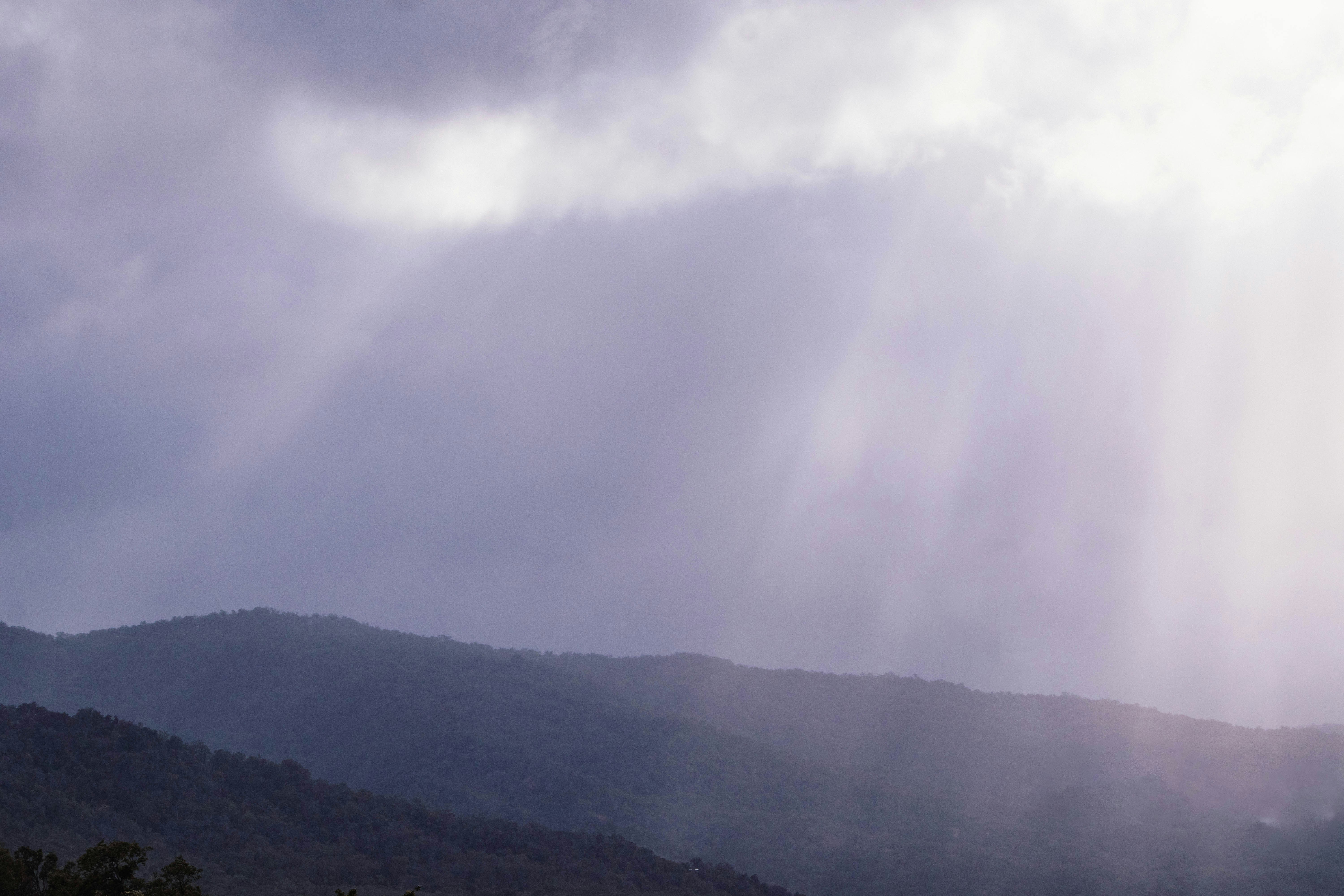 Sunbeams slip through a cloudy sky over distant ridge lines, capturing a tranquil landscape with soft light and layered hills.