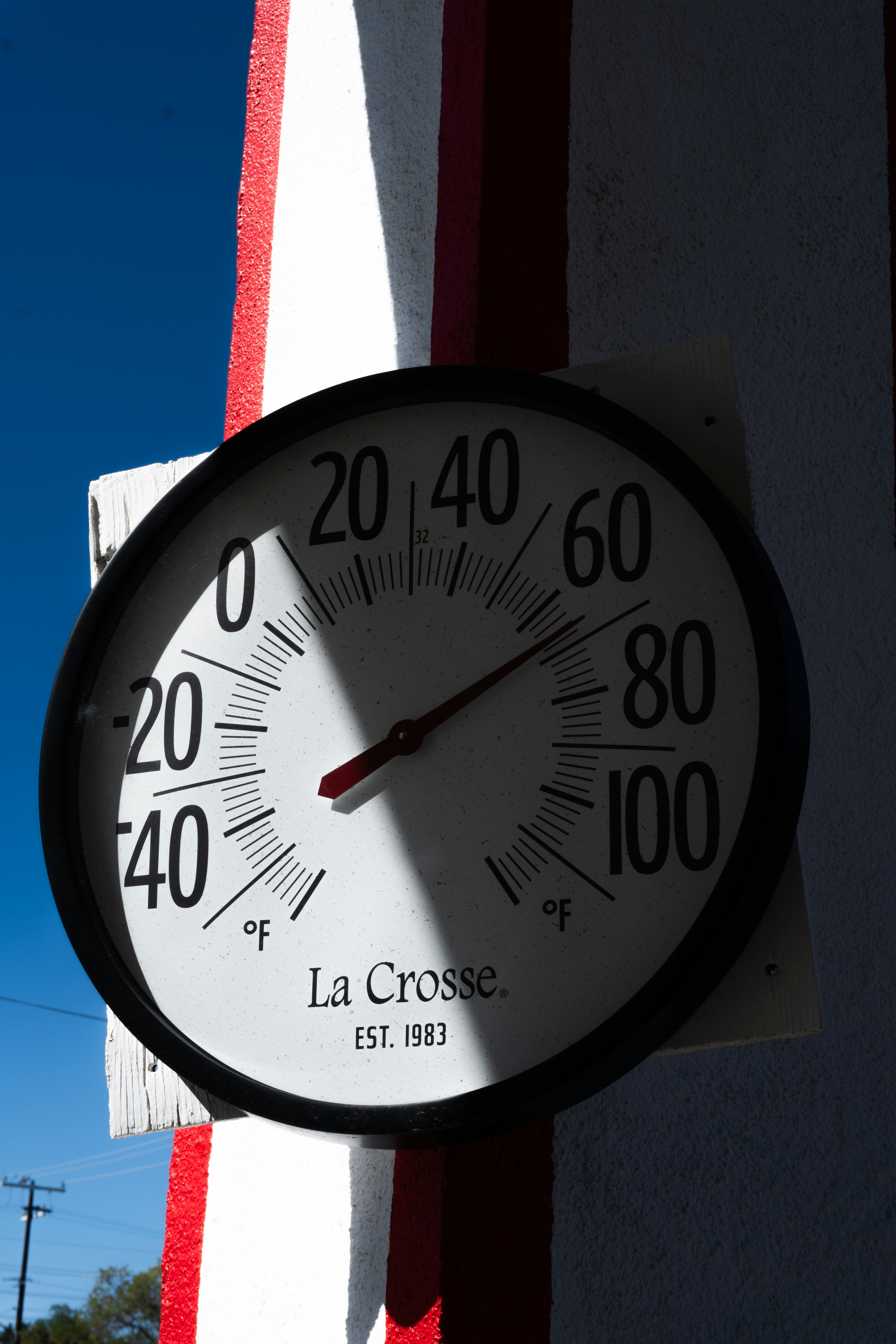Close-up of a round outdoor thermometer mounted on a white wall with red trim, showing a blue sky in the background and the brand 'La Crosse' at the bottom.