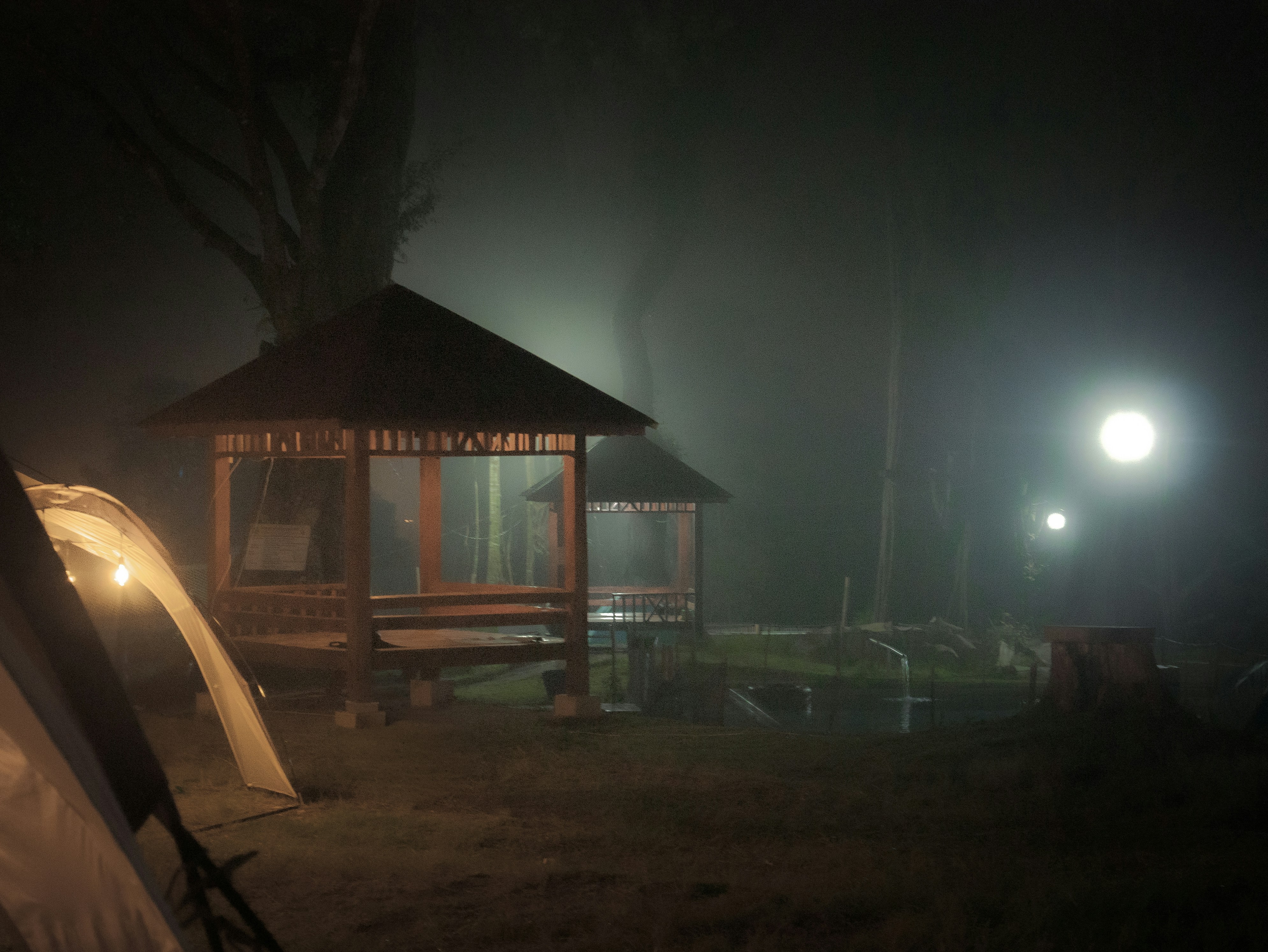 A misty nighttime campsite featuring wooden gazebos, tents, and glowing lights.