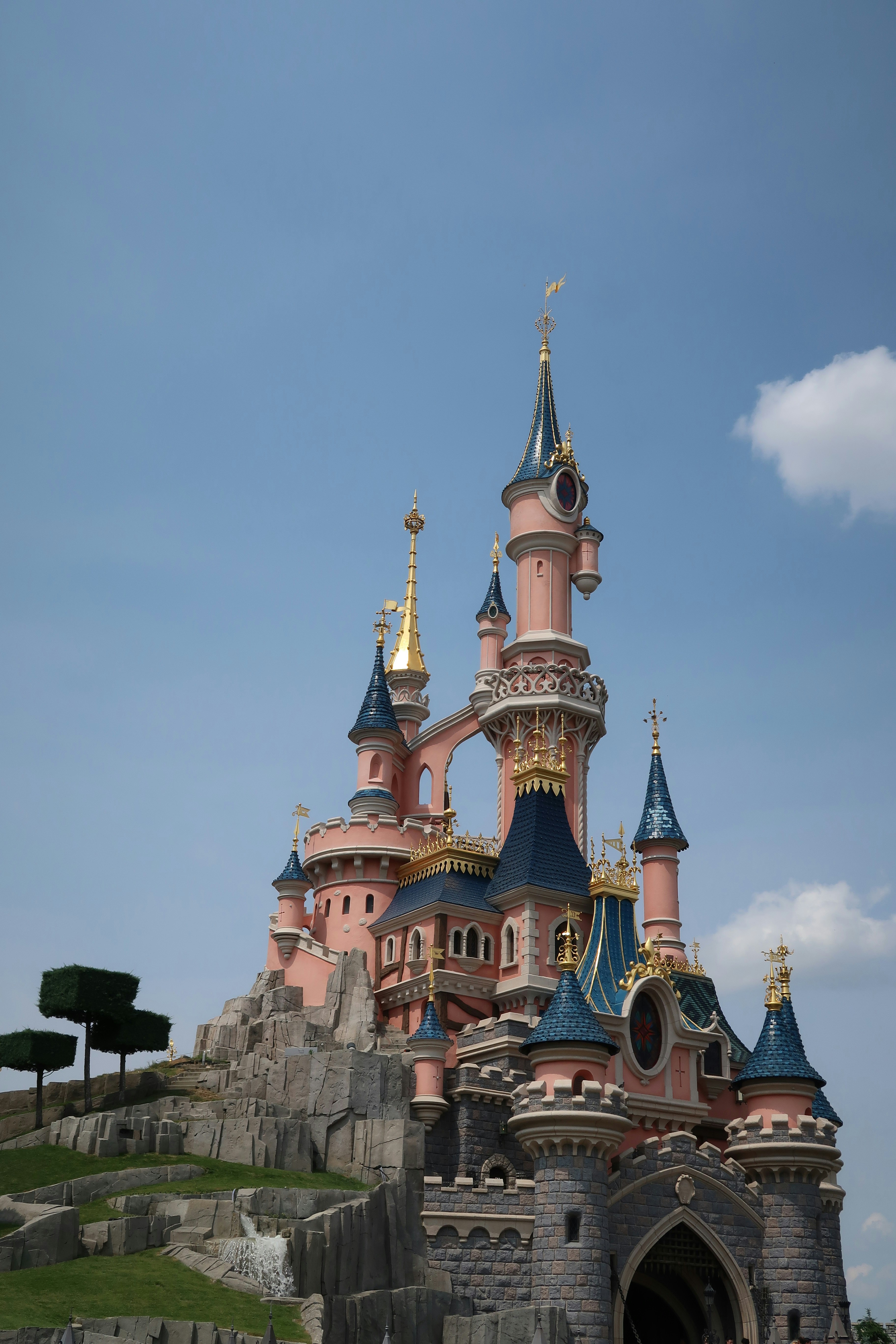 A pink-and-blue fairy-tale castle with gold accents rises from rocky terrain against a clear blue sky, capturing a whimsical theme park centerpiece.