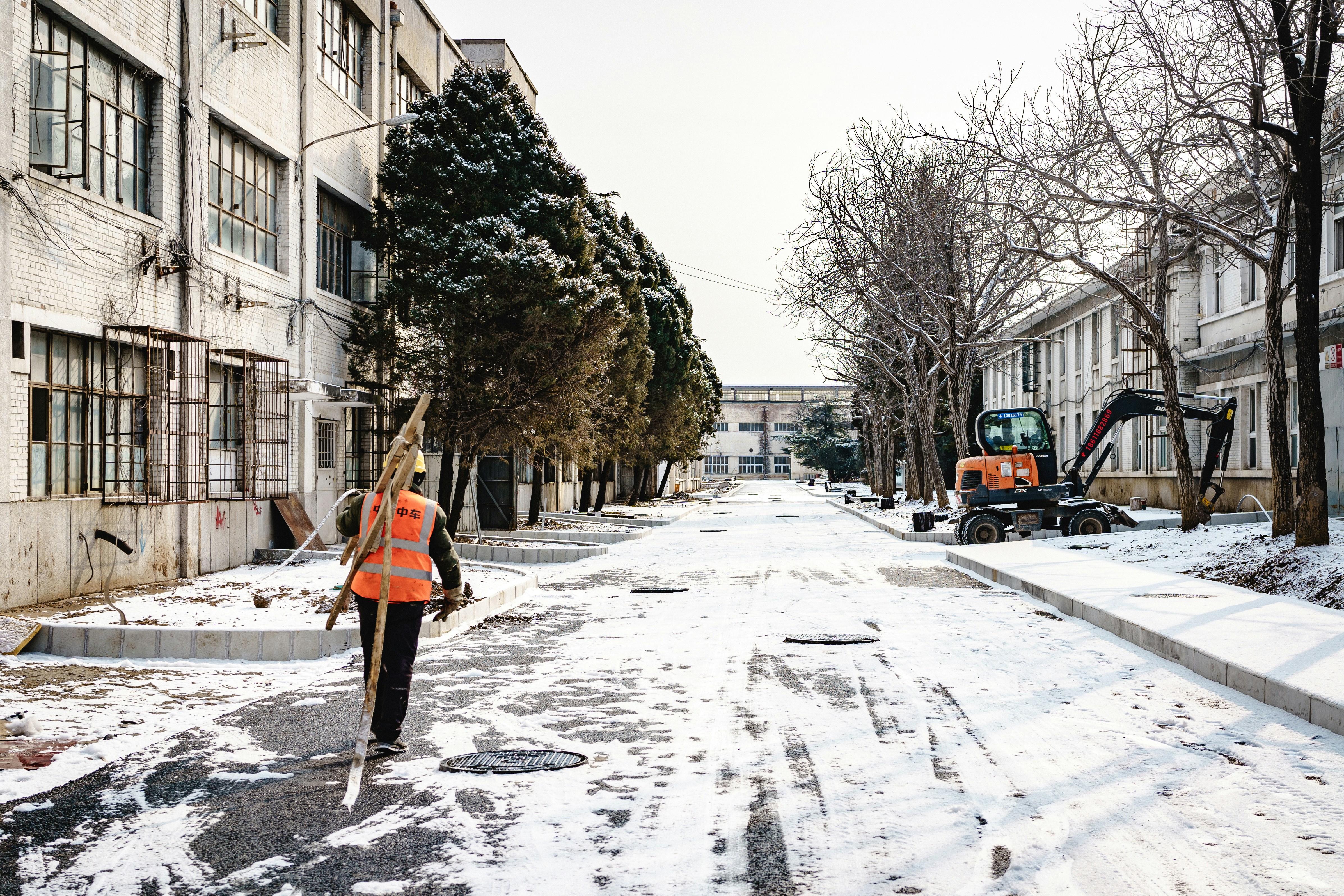 Winter Crewman in Neon Vest Guiding Snow-Clearing with Backhoe in an Idle Campus Lane