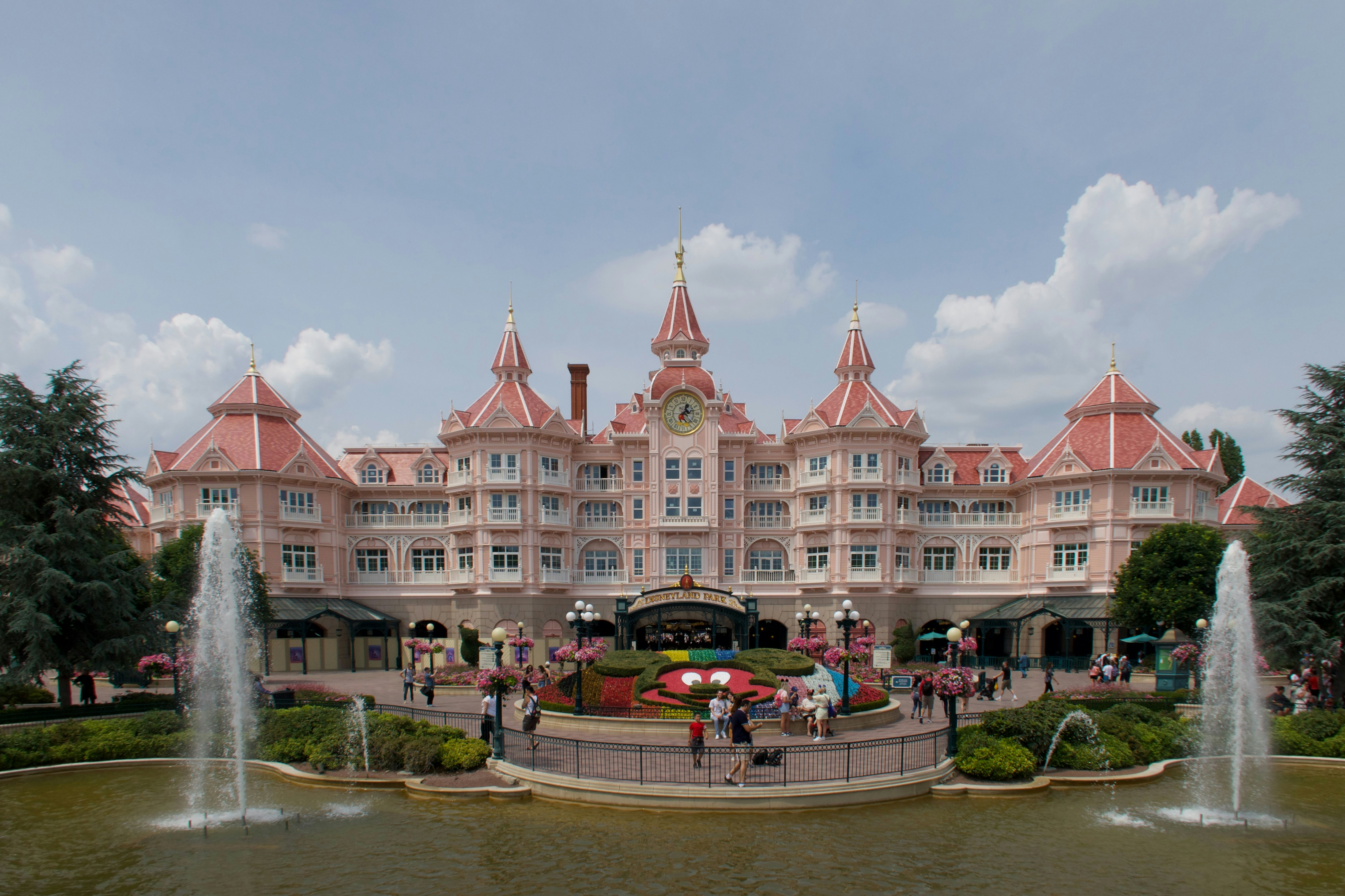 A grand pink castle-style hotel sits behind a manicured plaza with two arcing fountains and a circular pond, lush greenery, and visitors in the foreground.