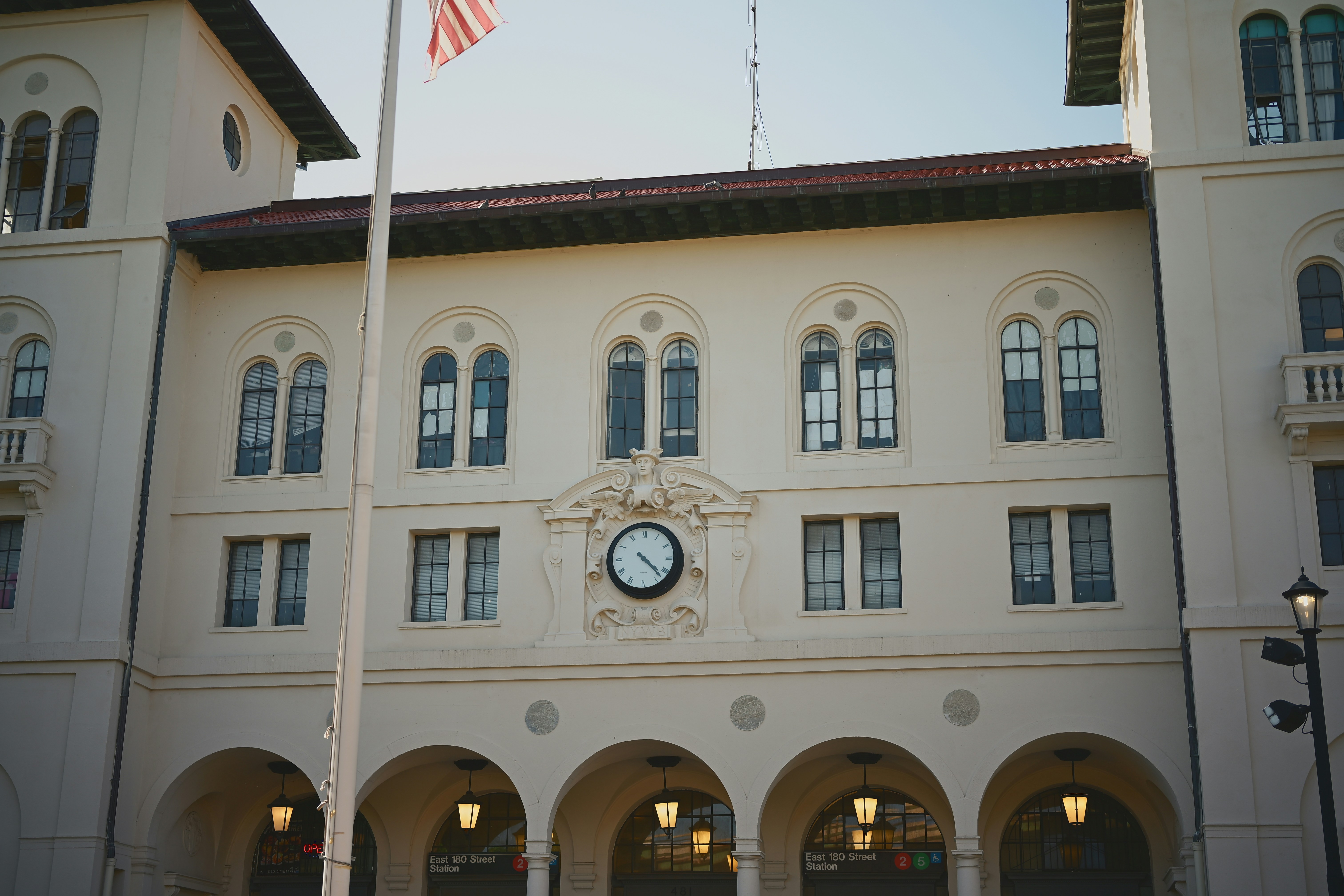 Classic façade with a central clock above arched colonnade and symmetrical windows, flagpole and warm lamps illuminating the arcade.