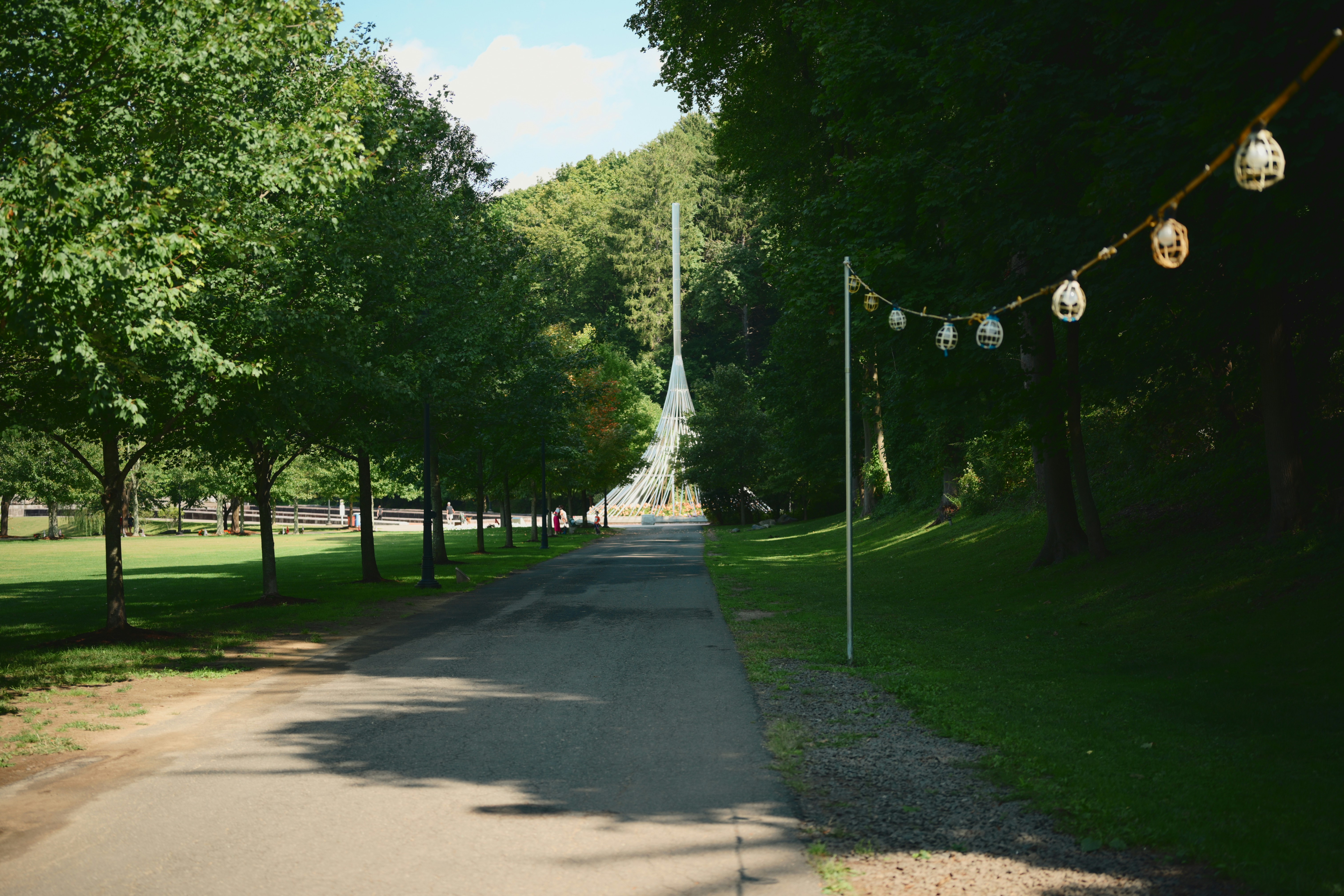 Tree-lined path with string lights leading to a white spire.
