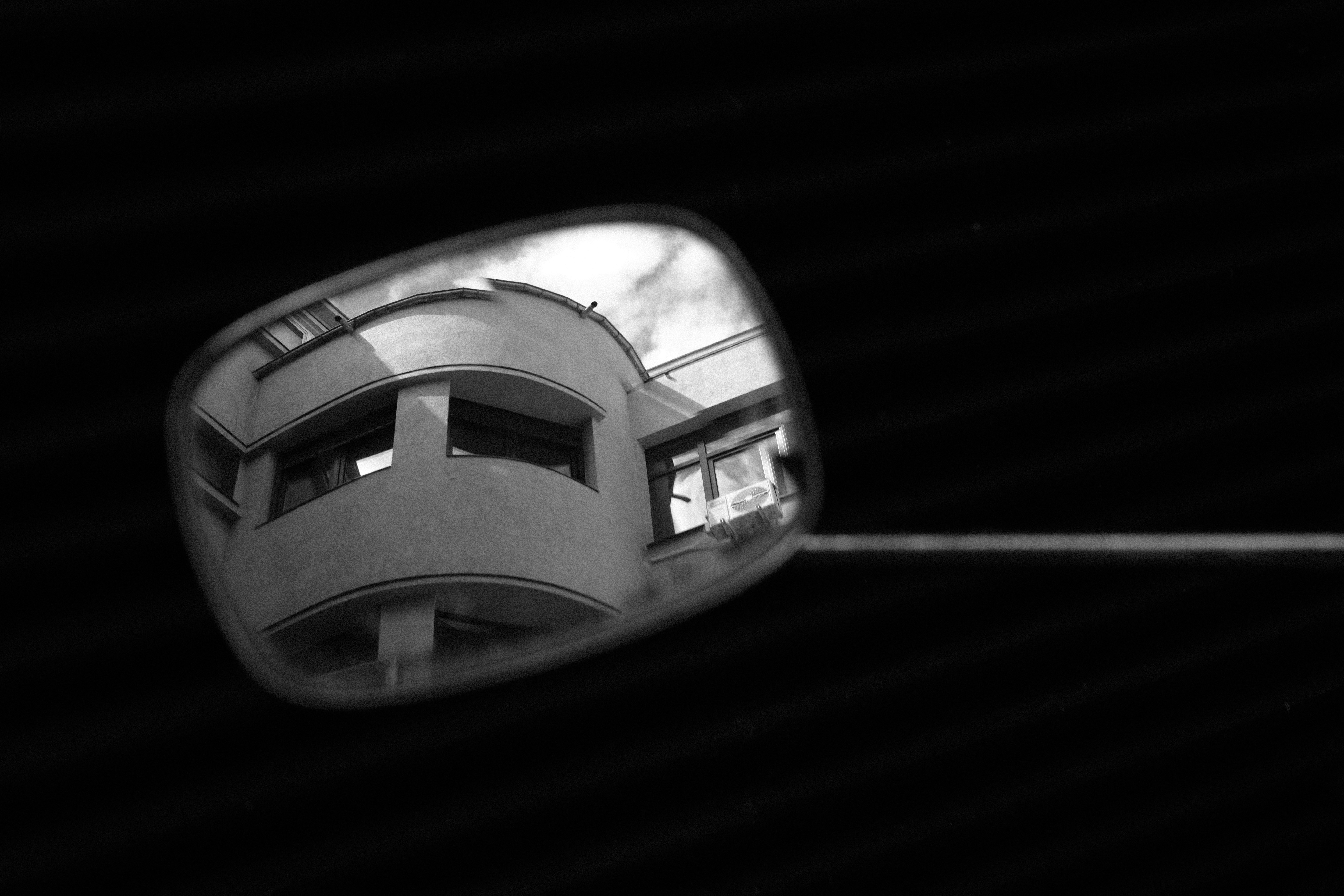 A grayscale reflection of a rounded building facade appears in a small, curved mirror, set against dark, horizontal ridges with a single wire crossing the frame.