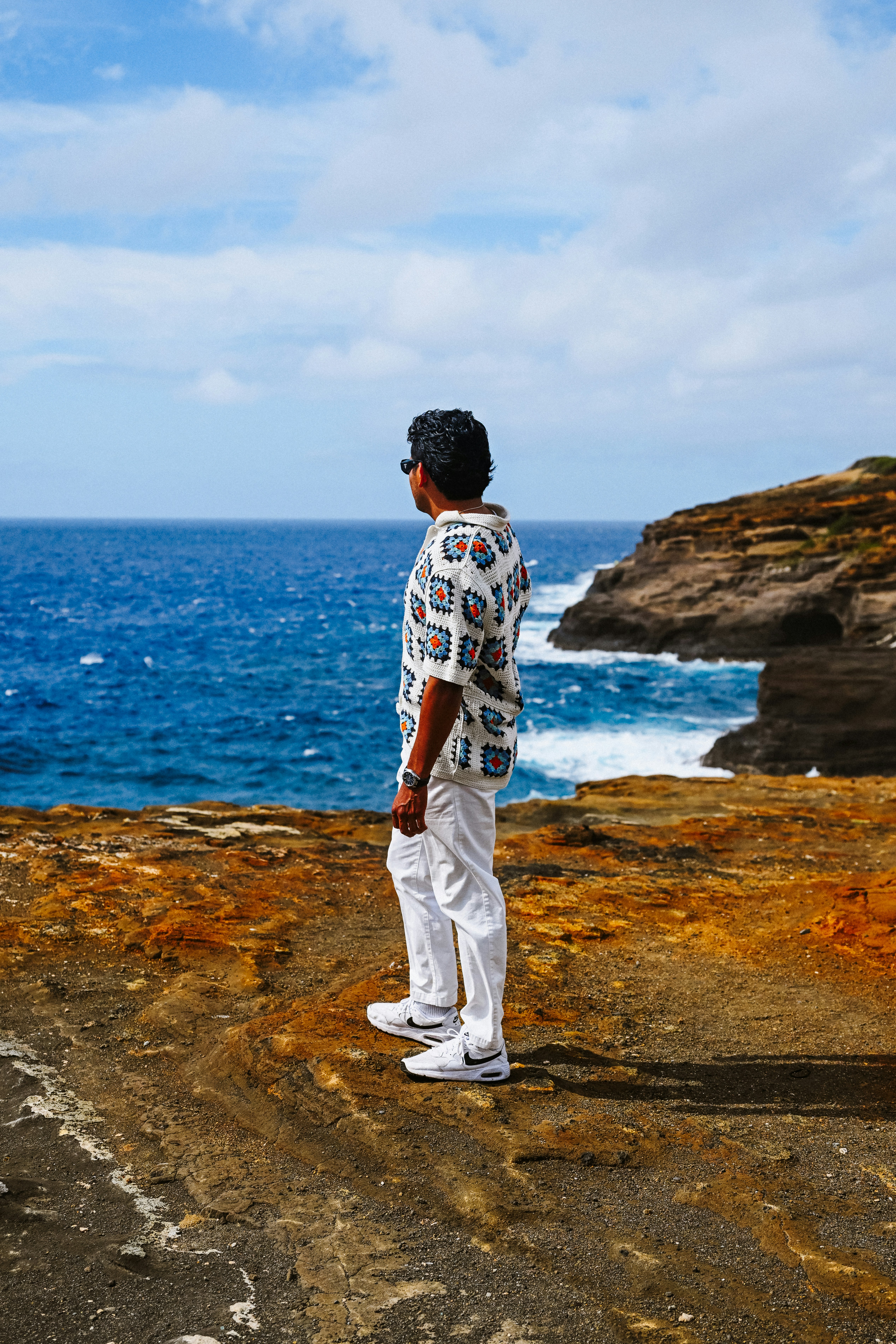 Man in white stands on cliff looking at blue ocean.